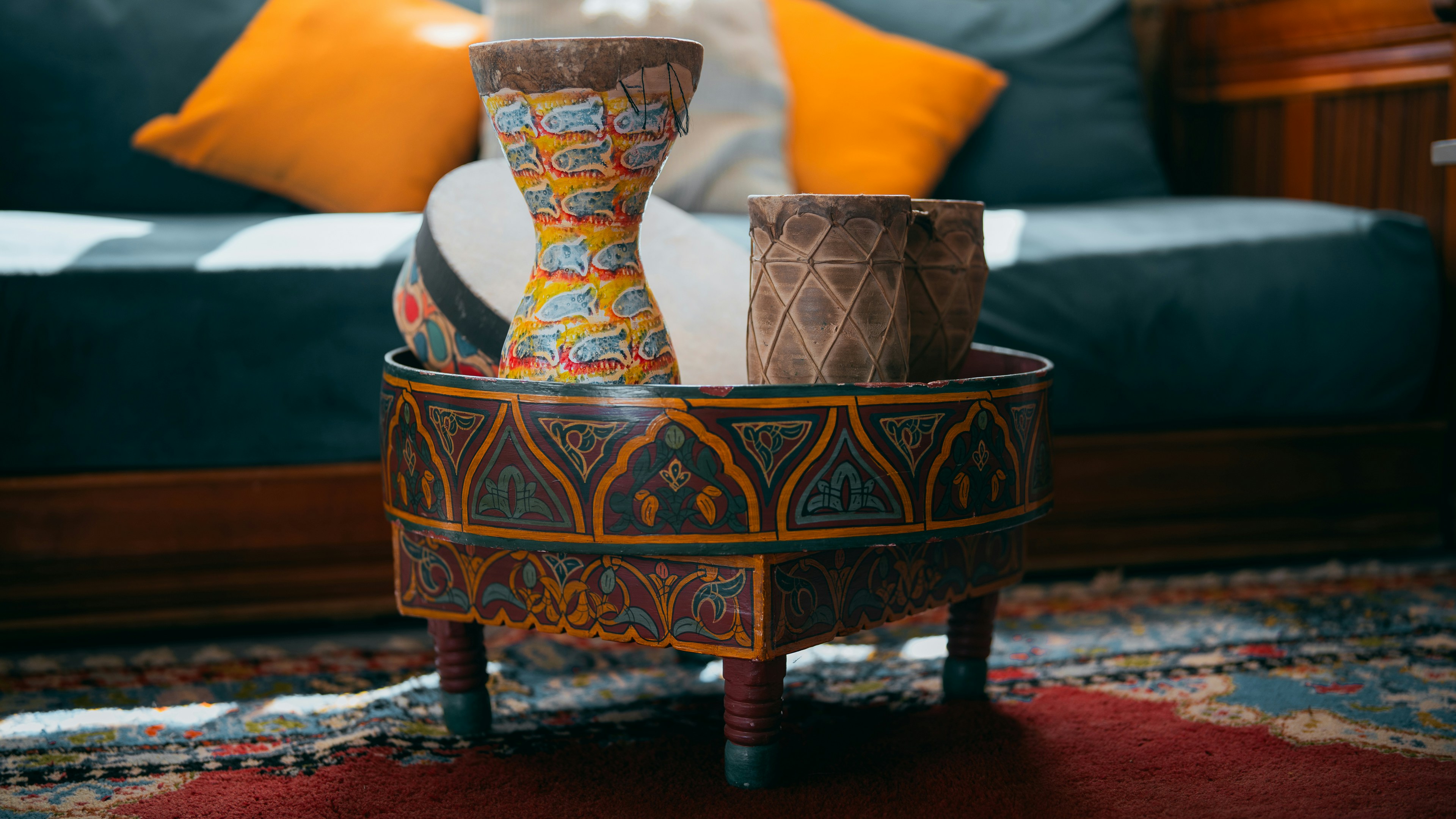 A richly patterned footstool with two decorative boots sits atop a carved wooden table, set against a colorful rug and a blue sofa with orange cushions in the background.