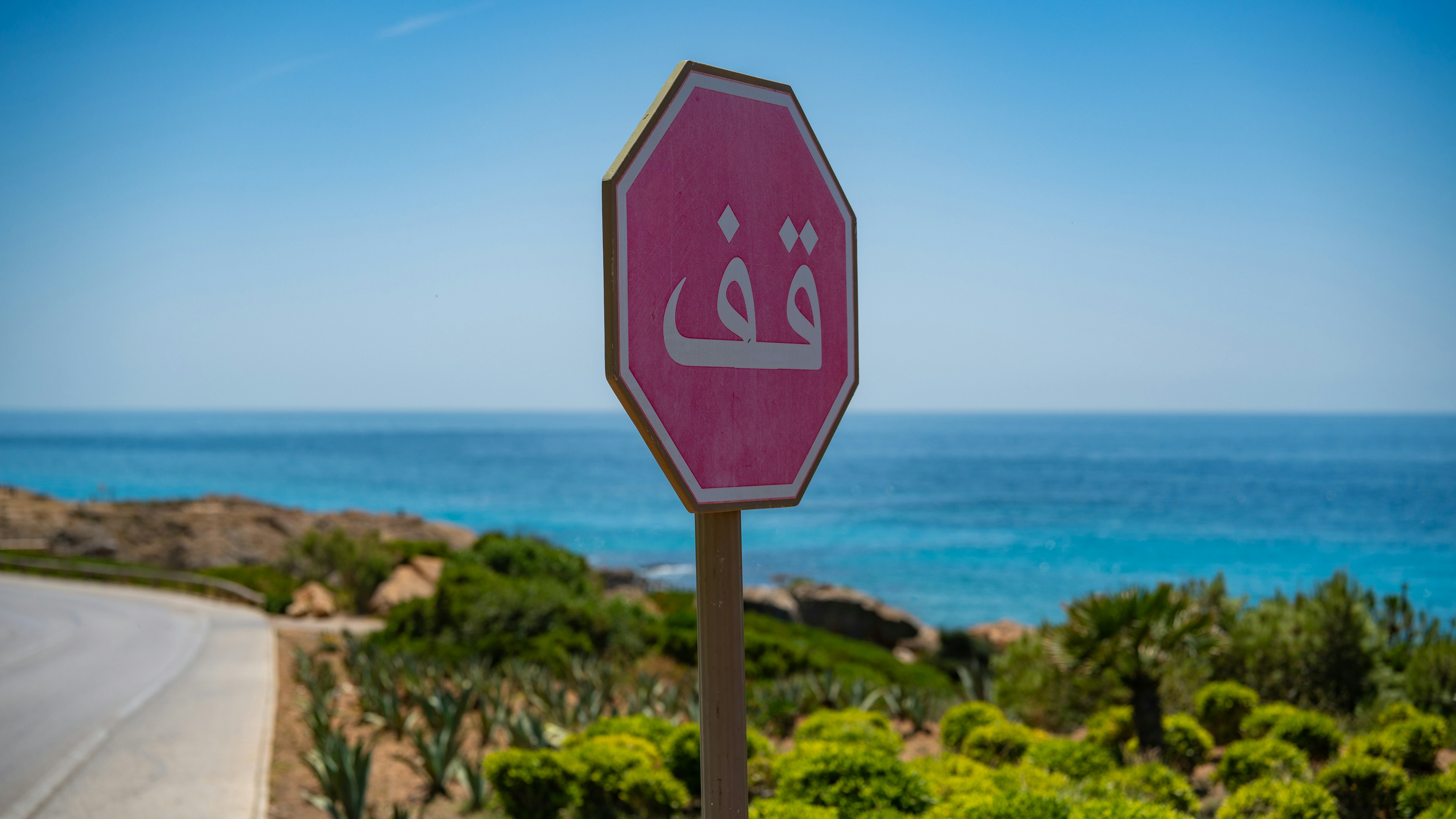 Pink stop sign with arabic text by a blue ocean.
