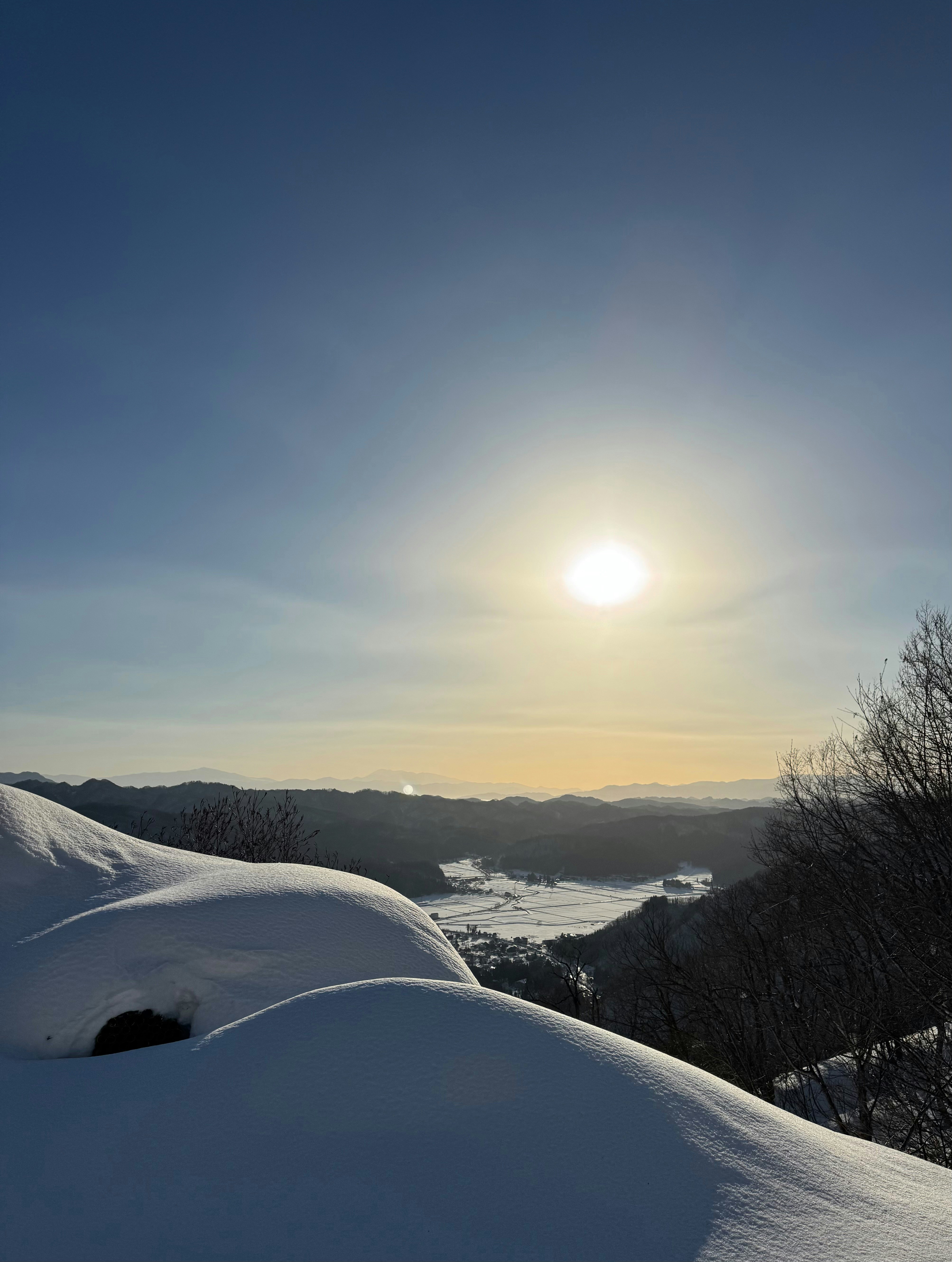 Deep, untouched snow fills the foreground in soft mounds, with a dark hollow carved at the base of one drift. Beyond, a small village sits in a white valley floor, flanked by bare trees on a steep hillside. Layers of mountain ranges recede into the distance in shades of blue and grey. The sun hangs high in a clear sky, casting a warm glow along the horizon and bathing the cold, quiet scene in gentle light.