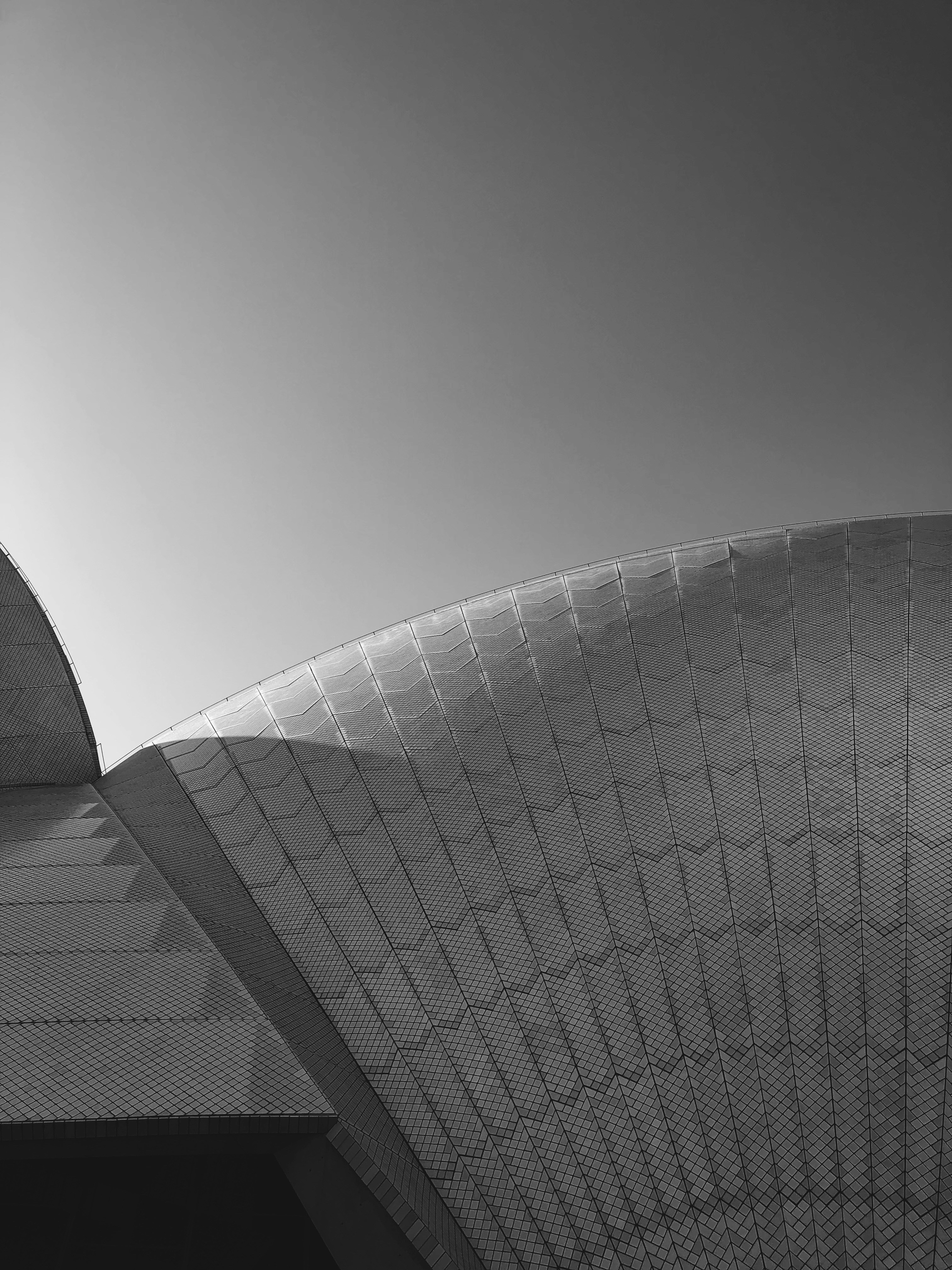 A black-and-white close-up of the Sydney Opera House roof shells, shot from below. Chevron-patterned tiles cover the curved surfaces in precise repeating rows, while a sweeping shadow cast by one sail onto another reveals the building’s sculptural depth. Directional light from the upper left picks out texture and form against a smooth, gradient sky. The composition is minimal and abstract — all geometry, contrast, and clean edges.