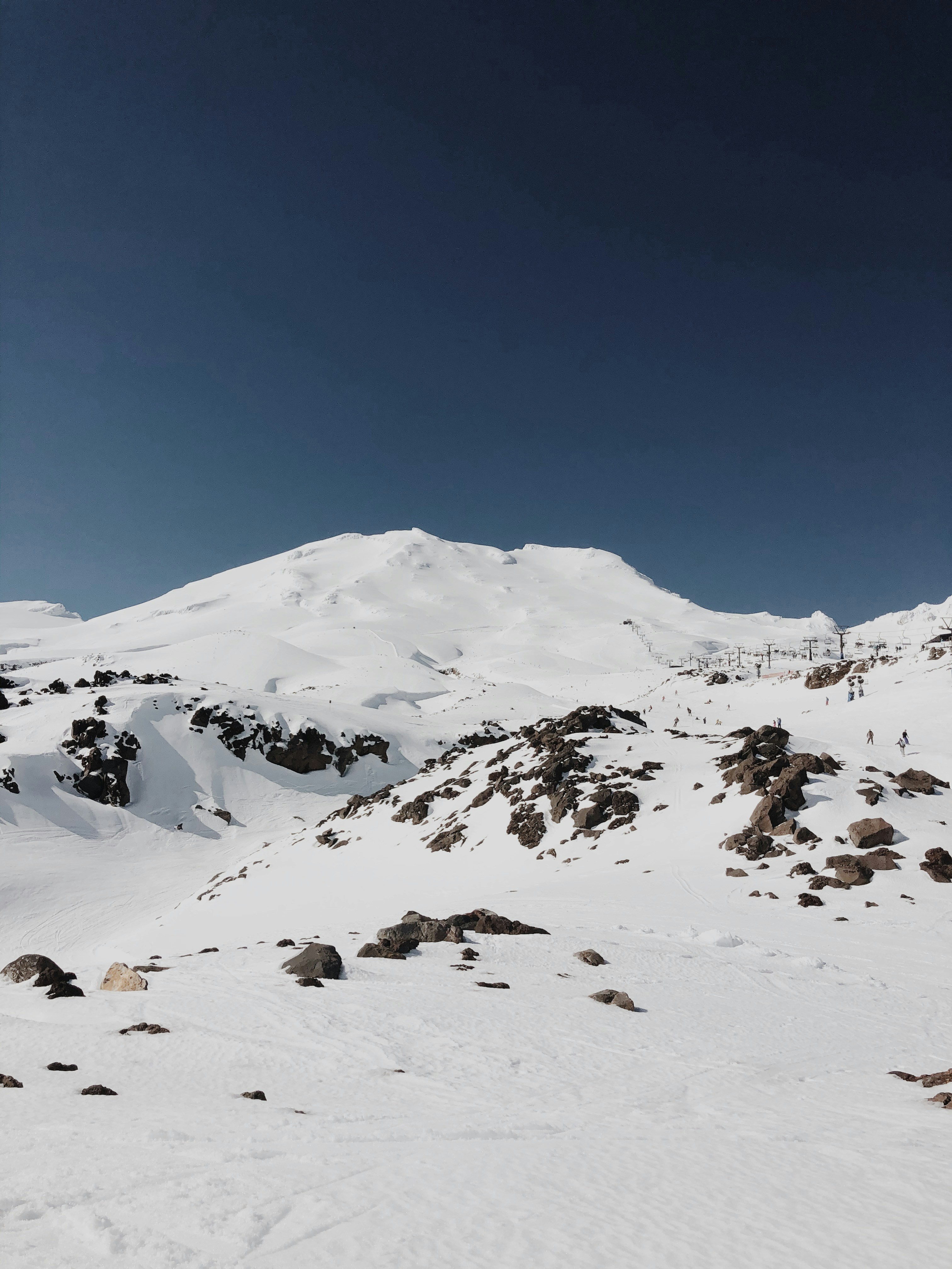 A smooth, snow-covered mountain peak rises under a deep blue sky, its contours softened by a thick blanket of white. Ski lift poles trace a line along the lower slopes where tiny figures carve their way down. In the foreground, dark rocks break through the snow on a gentle slope, casting crisp shadows in the bright overhead sun. The air looks cold and clear — a perfect bluebird day on the mountain.