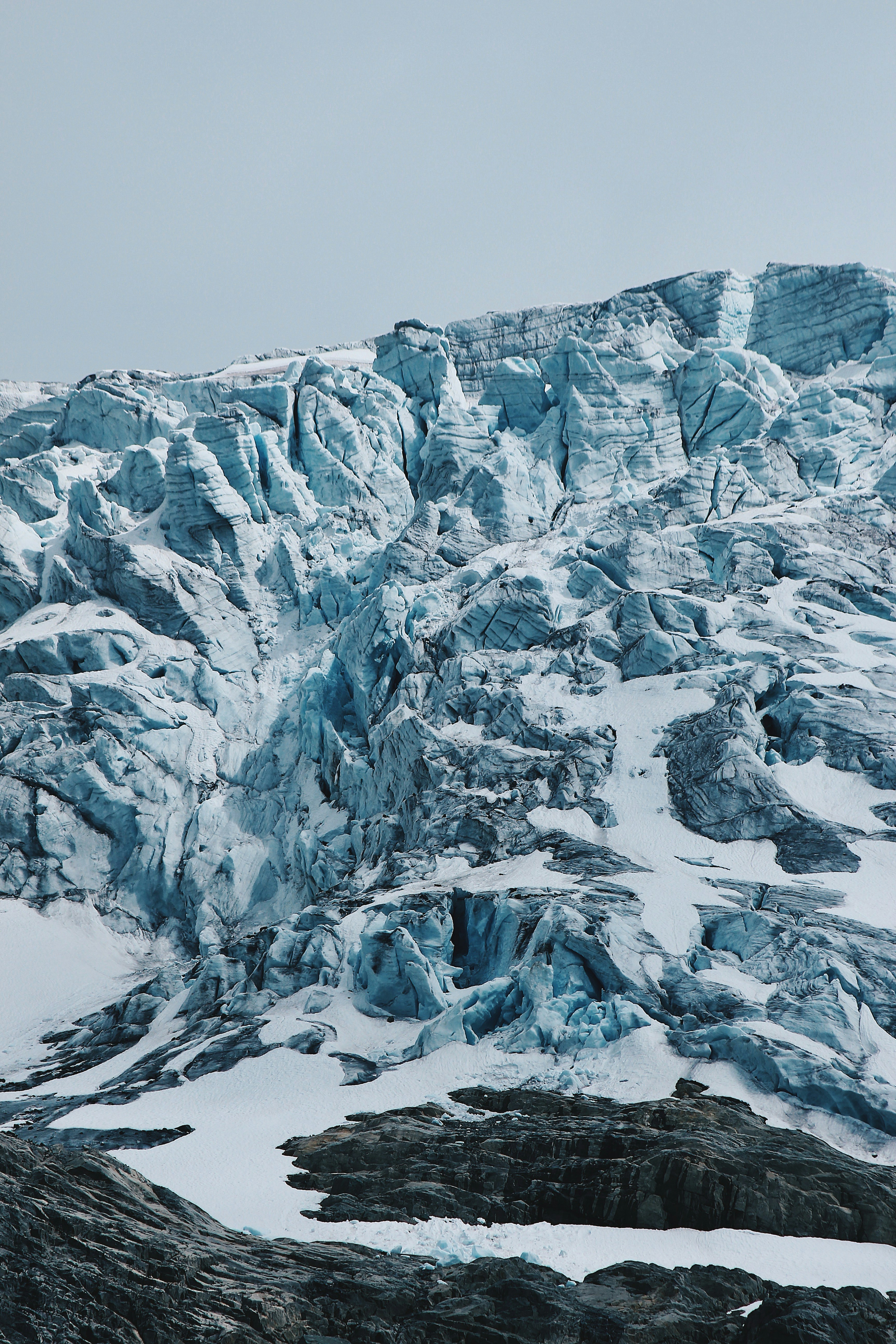 A towering glacier wall fills the frame, its ice a brilliant pale blue deepening to rich tones inside crevasses and layered fractures. Sharp ridges and pinnacles rise from the frozen mass, sculpted by time and pressure. Patches of white snow settle into the hollows, and dark rocky outcrops emerge at the base. Shot from a low angle under a flat, overcast sky, the scene is raw and immense — no vegetation, no people, just ice and stone.