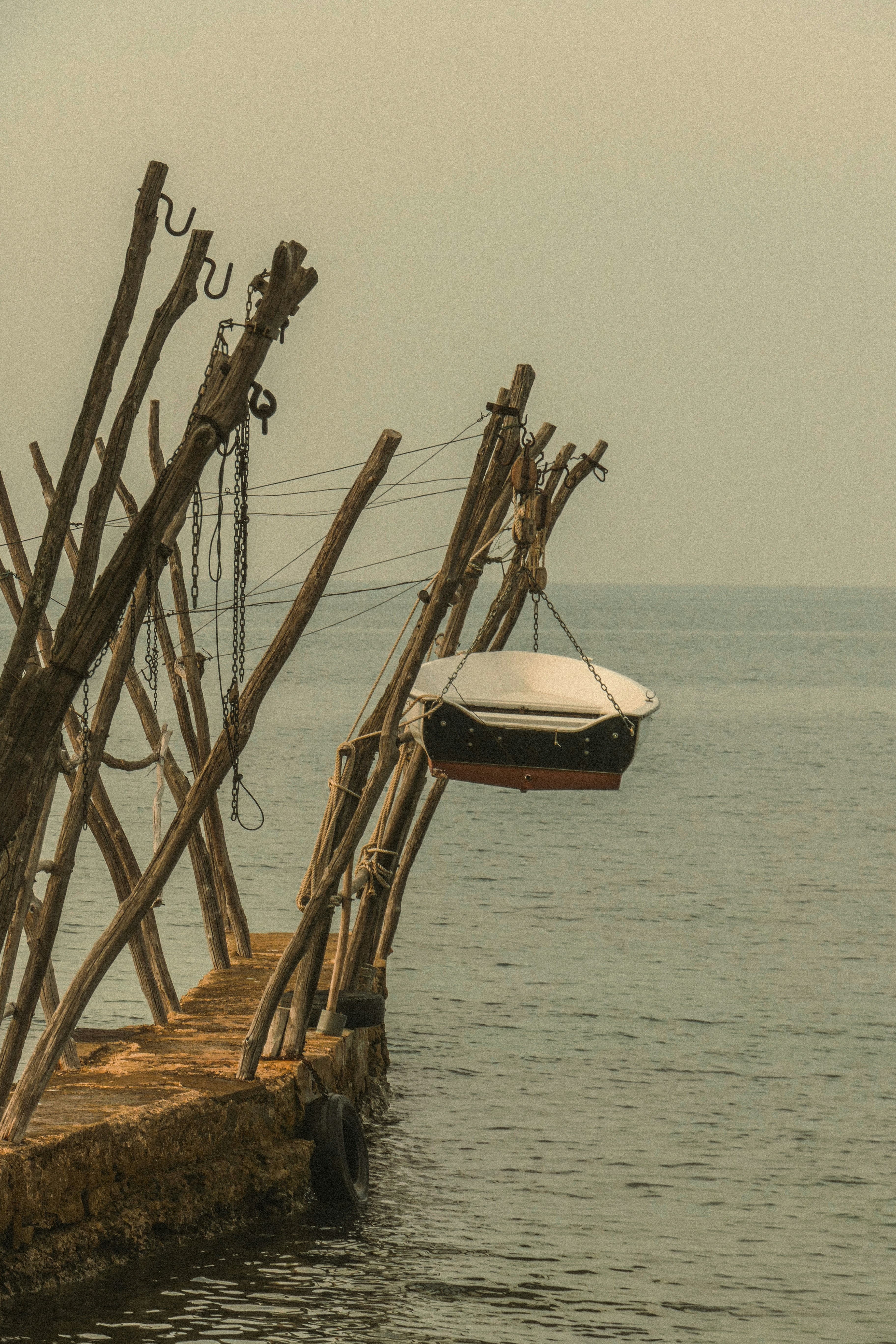 Weathered wooden posts form a rustic pier with a single hanging boat over calm water, capturing a quiet seascape with muted tones.