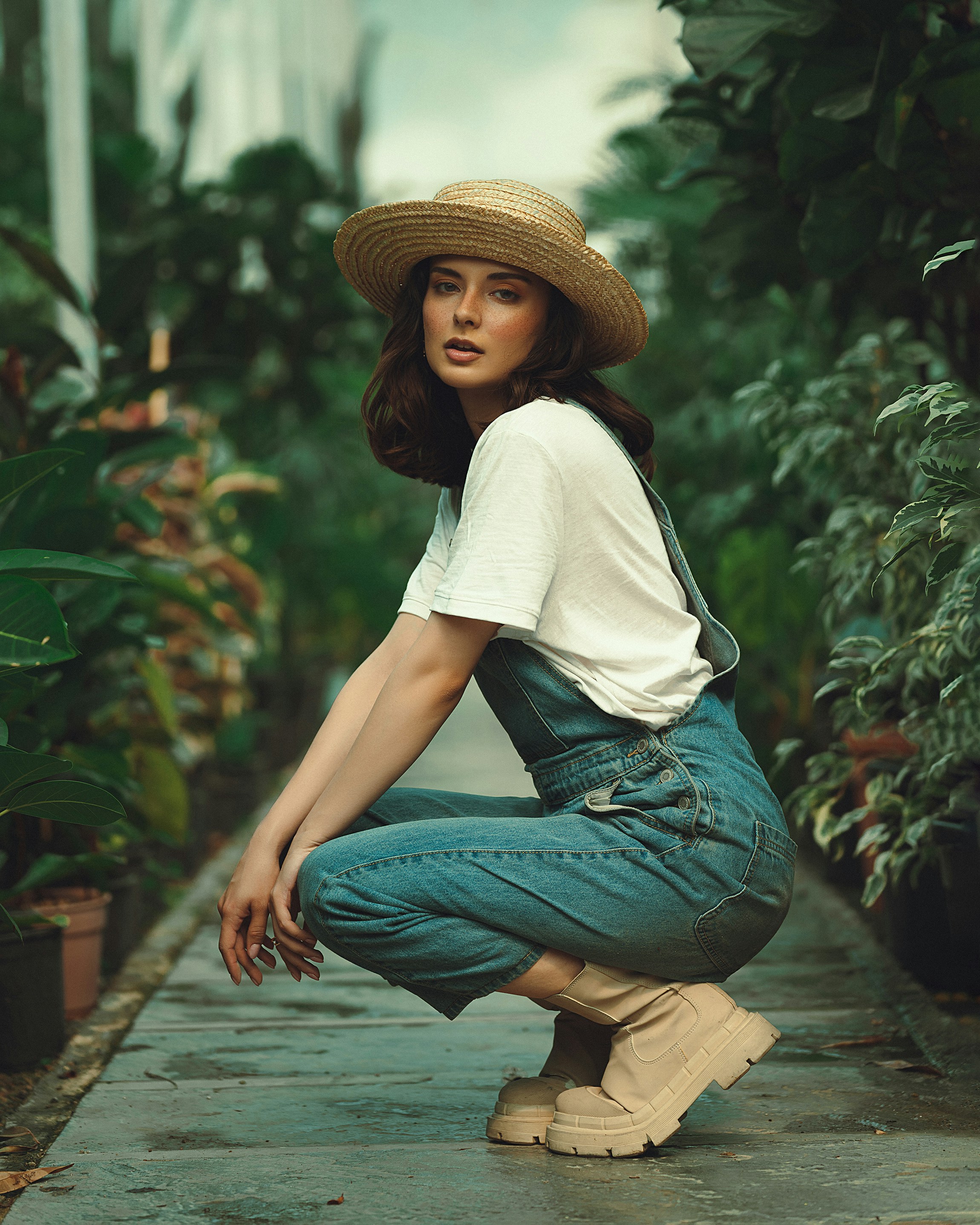 Woman in straw hat and overalls crouching among green plants.