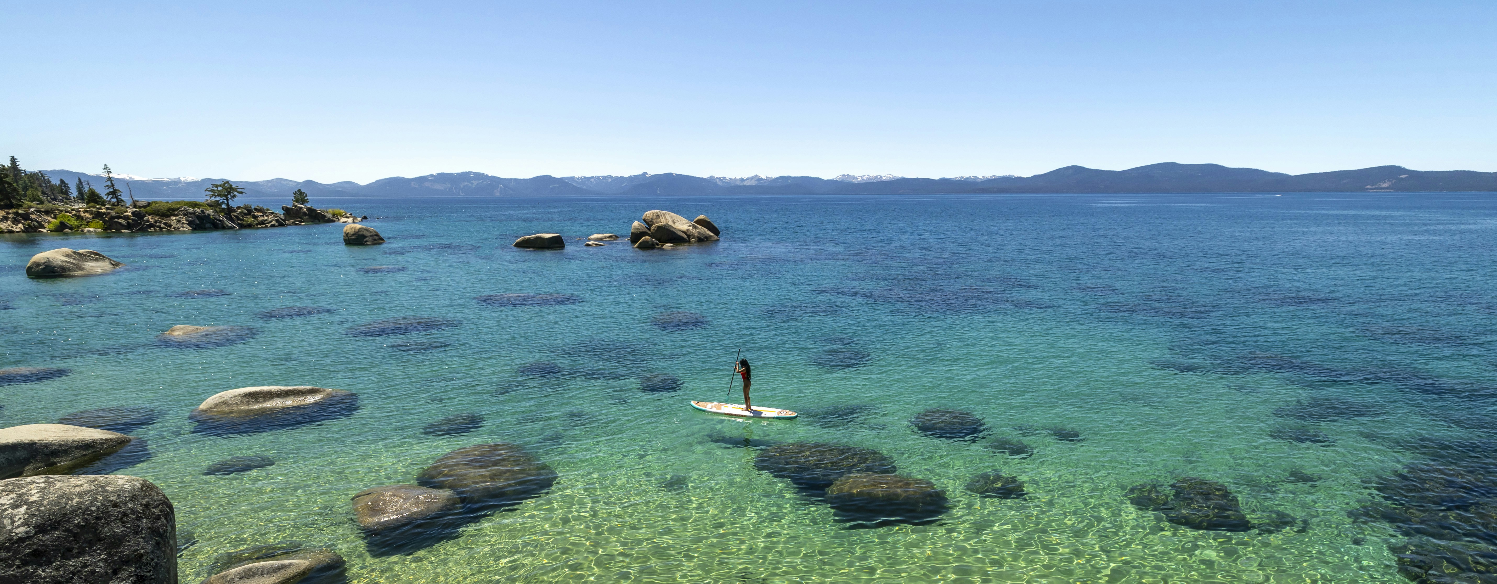 Paddleboarder glides across clear turquoise lake water with visible rocks.