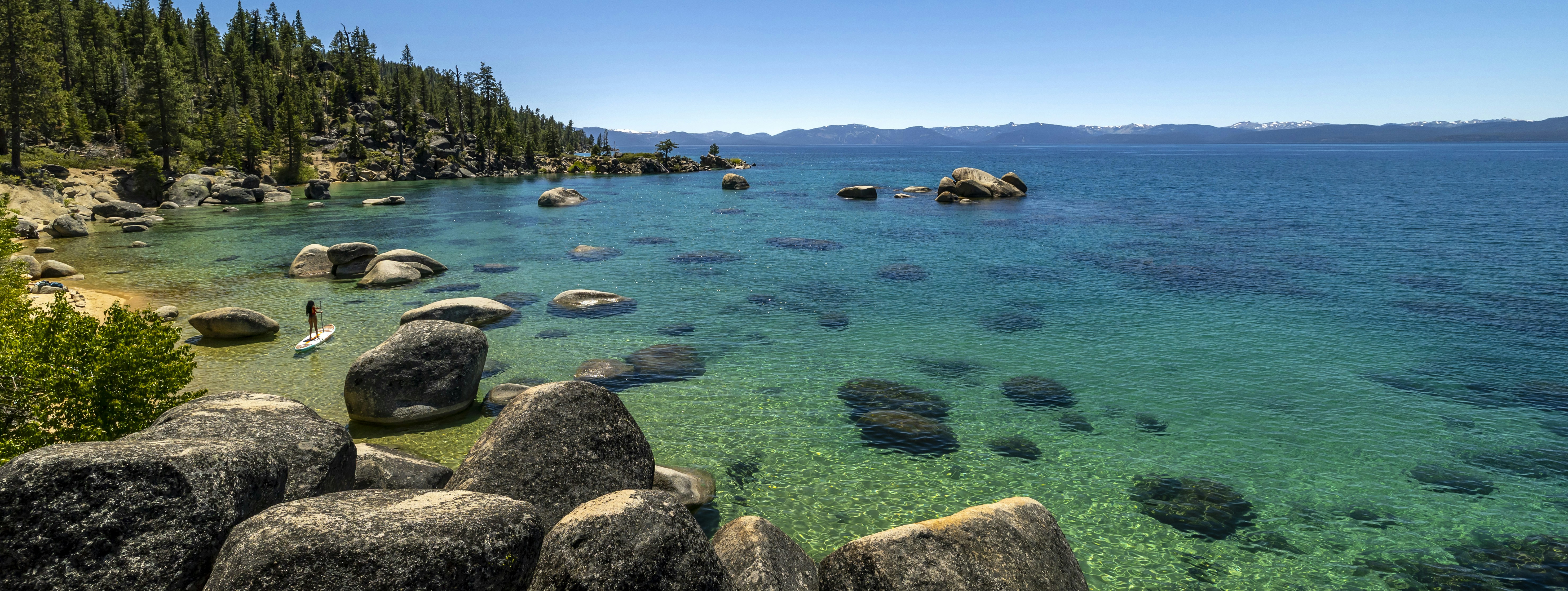 Paddleboarder on clear turquoise lake surrounded by rocks and trees.