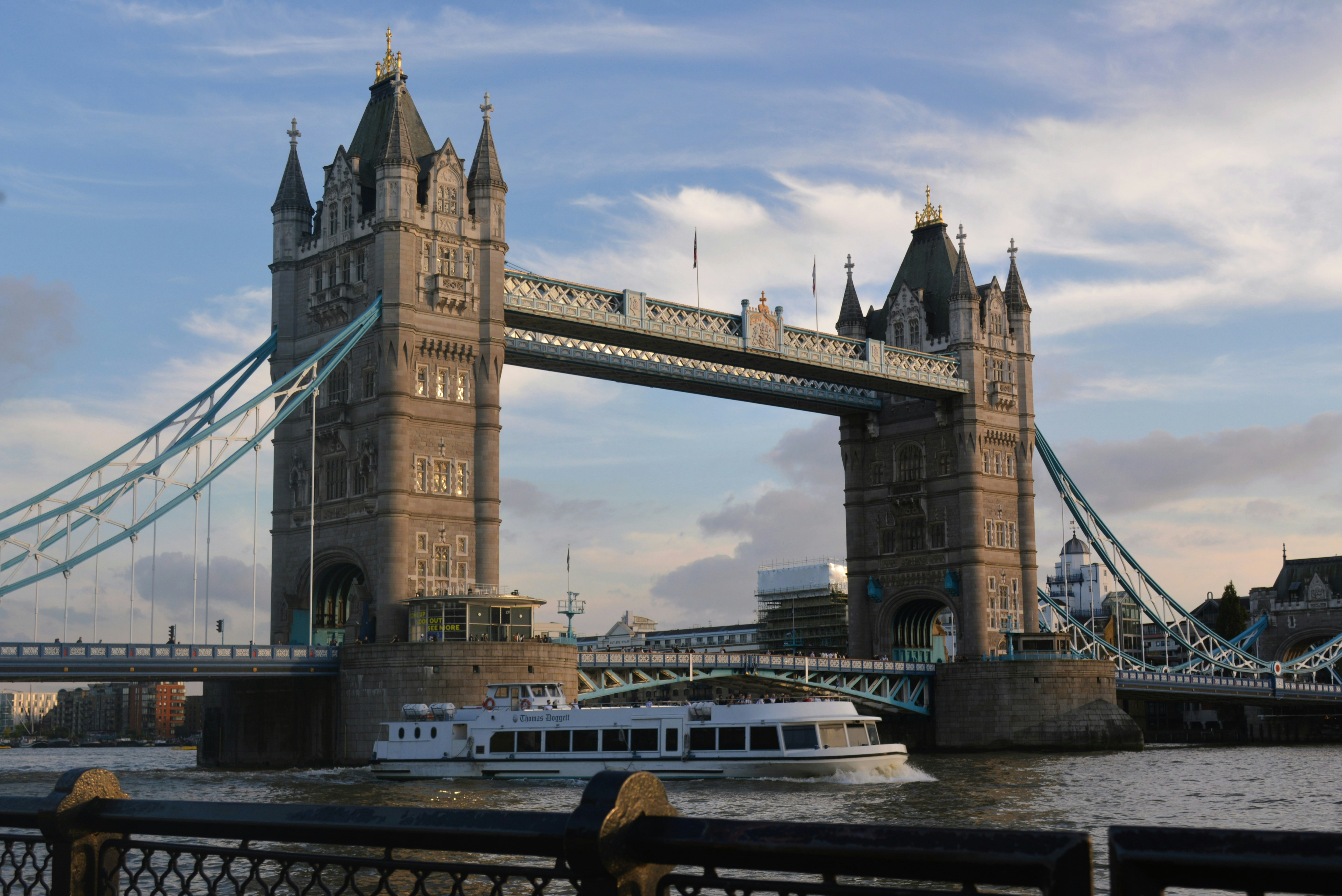 A classic, majestic view of Tower Bridge standing proudly over the River Thames in London, United Kingdom. The iconic Victorian Gothic towers and high-level walkways are beautifully illuminated by the warm, soft light of a late afternoon sun. In the foreground, a white double-decker river cruise boat glides across the water, passing beneath the bridge. Set against a soft blue sky filled with wispy clouds, this picture-perfect scene captures the timeless architectural grandeur, historic charm, and active maritime life of one of the world's most famous landmarks.