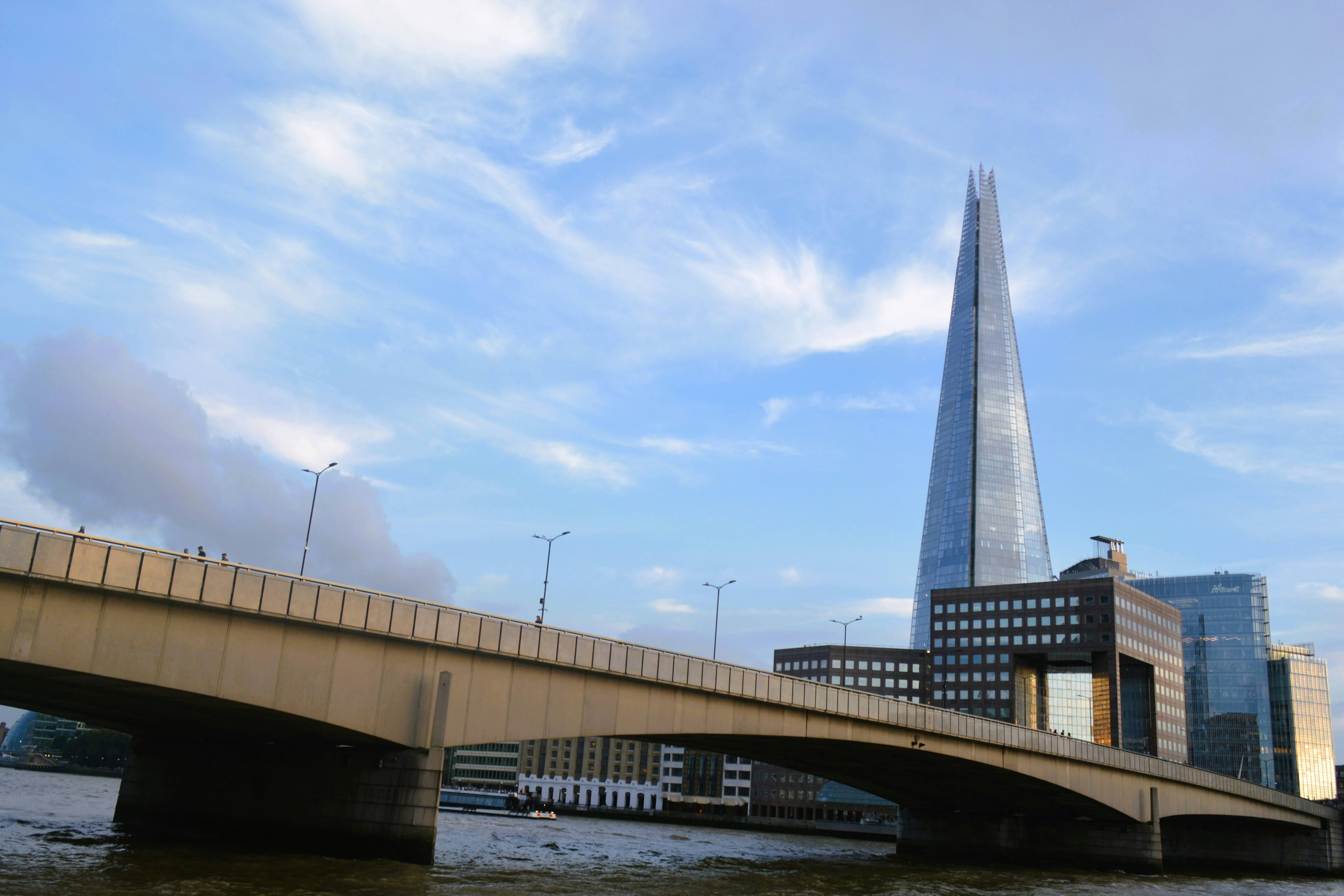 A striking perspective of London Bridge stretching across the River Thames in London, United Kingdom. In the background, the iconic, jagged glass spire of The Shard rises majestically into a beautiful blue sky filled with wispy, sweeping clouds. The modern corporate architecture surrounding London Bridge Station frames the base of the skyscraper, while the clean, minimalist lines of the concrete bridge draw the eye across the frame. This composition captures a crisp, bright day showcasing London's famous transit links and bold, contemporary architectural landmarks.