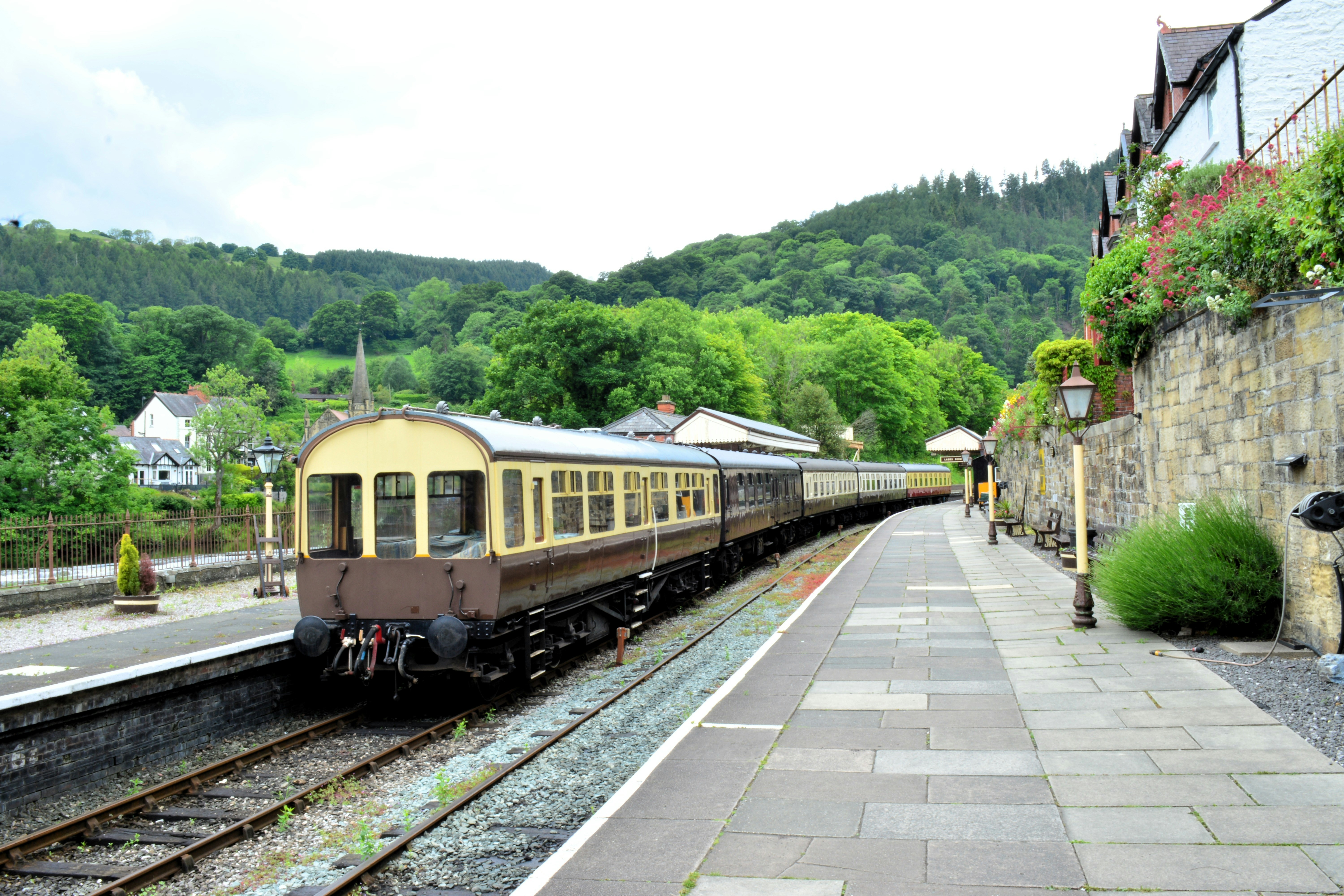 A charming, nostalgic scene at the historic Llangollen Railway Station in Denbighshire, Wales. A vintage steam or diesel heritage train with classic chocolate and cream carriages sits stationary alongside a long, curved stone platform. The right side is lined with a rustic stone wall adorned with colorful blooming flowers and traditional lampposts, while the left opens up to a view of a church spire and the lush, green wooded hills of the Dee Valley. This beautiful composition perfectly captures the timeless allure of British heritage rail travel on a peaceful, overcast day.