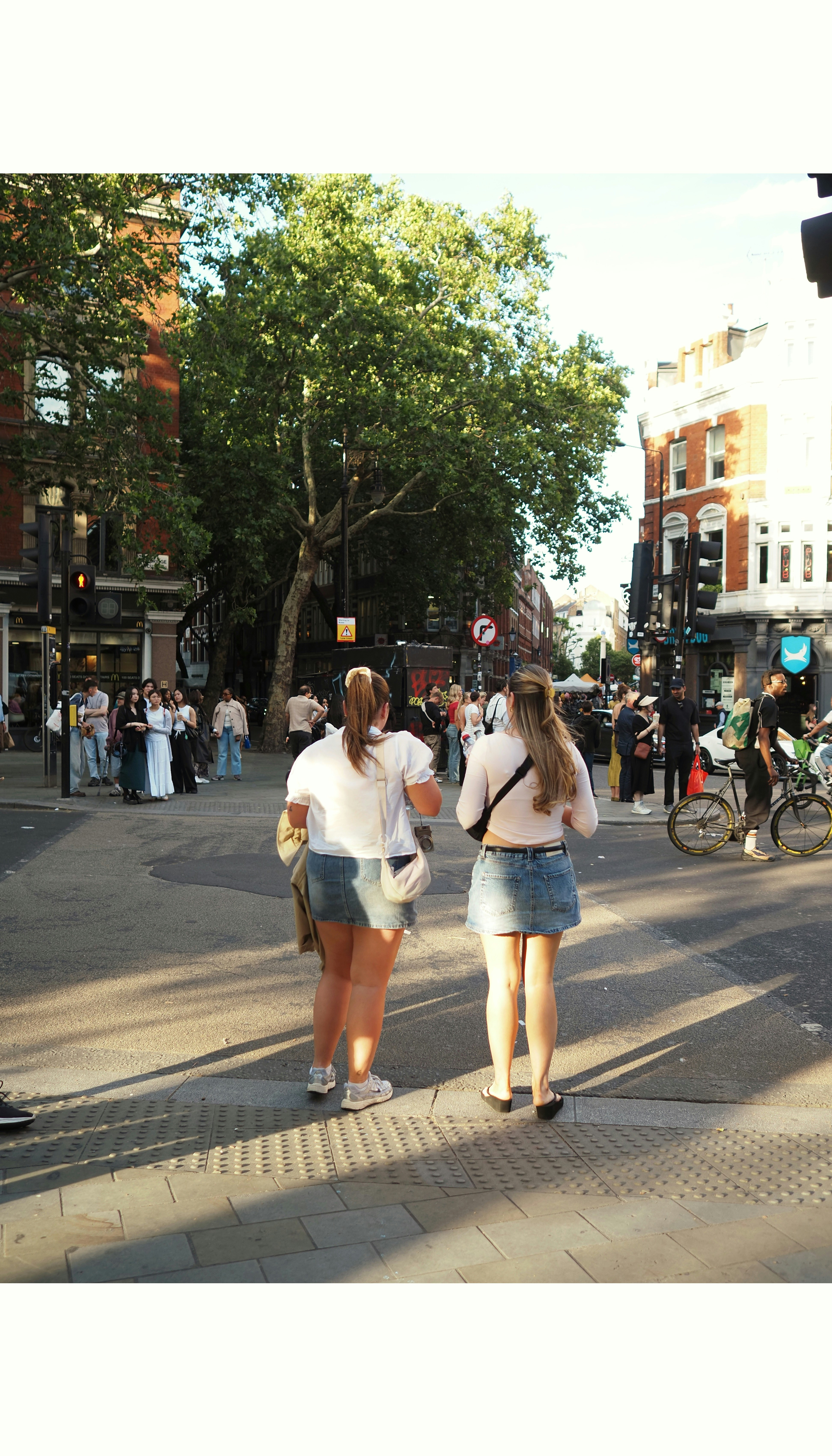 A warm summer evening at a busy London crossroads. Two friends pause at the curb as the city moves around them, bathed in golden light and long shadows. Street life, conversation, and the energy of Central London come together in a fleeting moment that feels uniquely British.