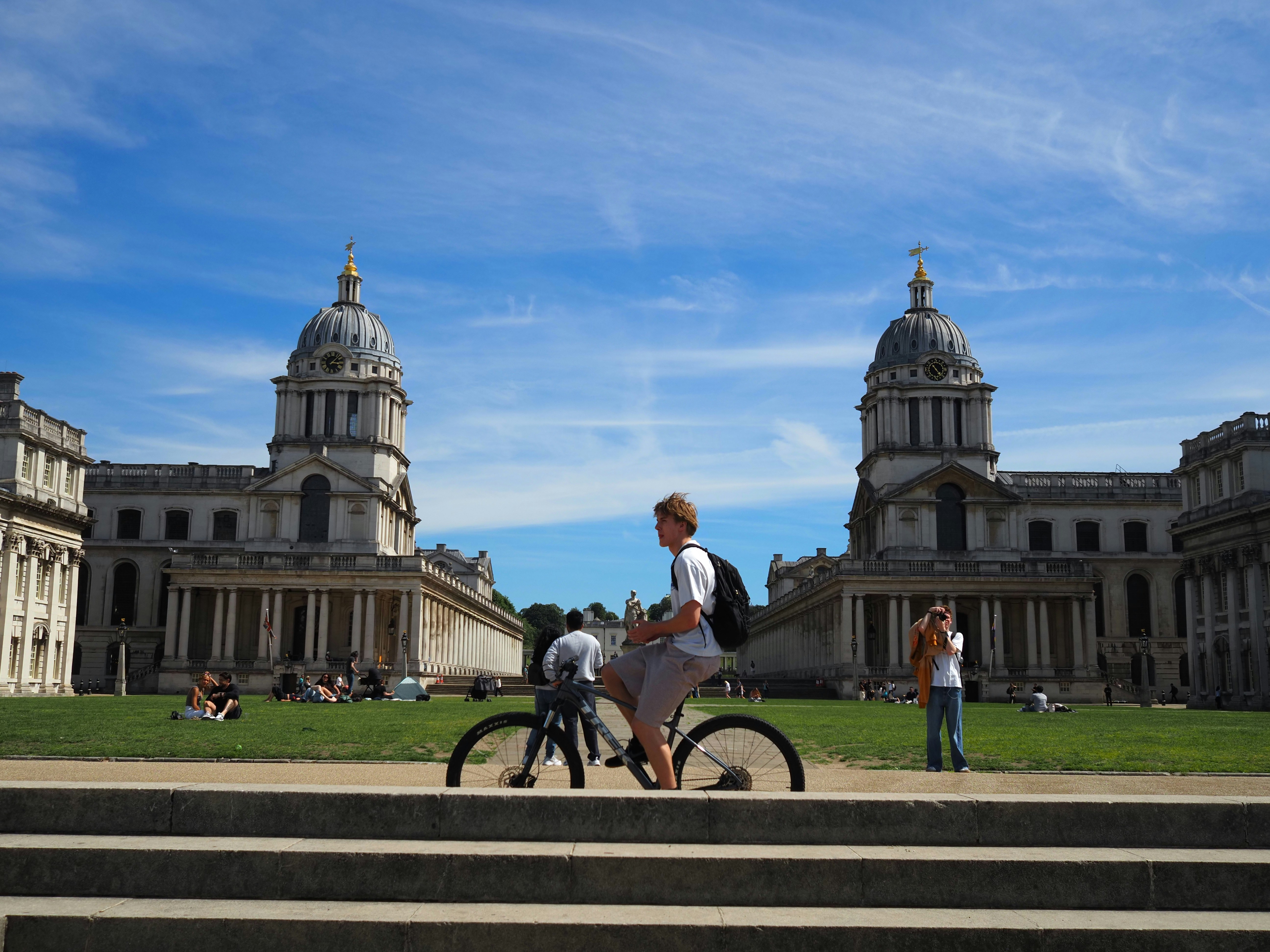 A cyclist passes through the heart of Greenwich as visitors relax on the lawns beneath the twin domes of the Old Royal Naval College. Framed by sweeping summer skies and classical architecture, the scene captures the calm pace of a perfect London afternoon.