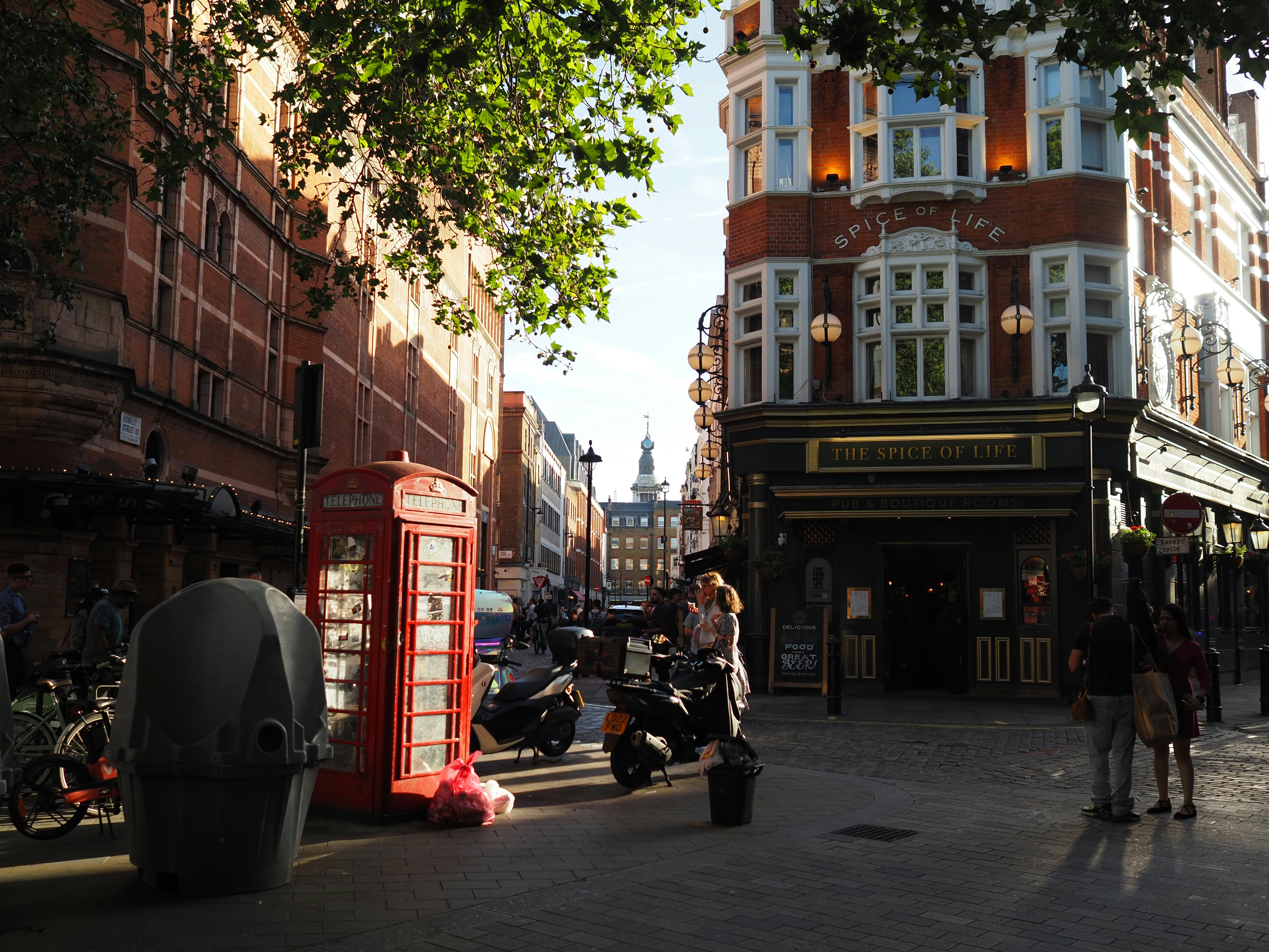 A classic London scene bathed in late afternoon light. A red telephone box stands beneath leafy trees, framed by historic brick buildings and the lively streets of Soho. The warm sunlight, long shadows, and timeless architecture capture the charm of a summer evening in the heart of the city.