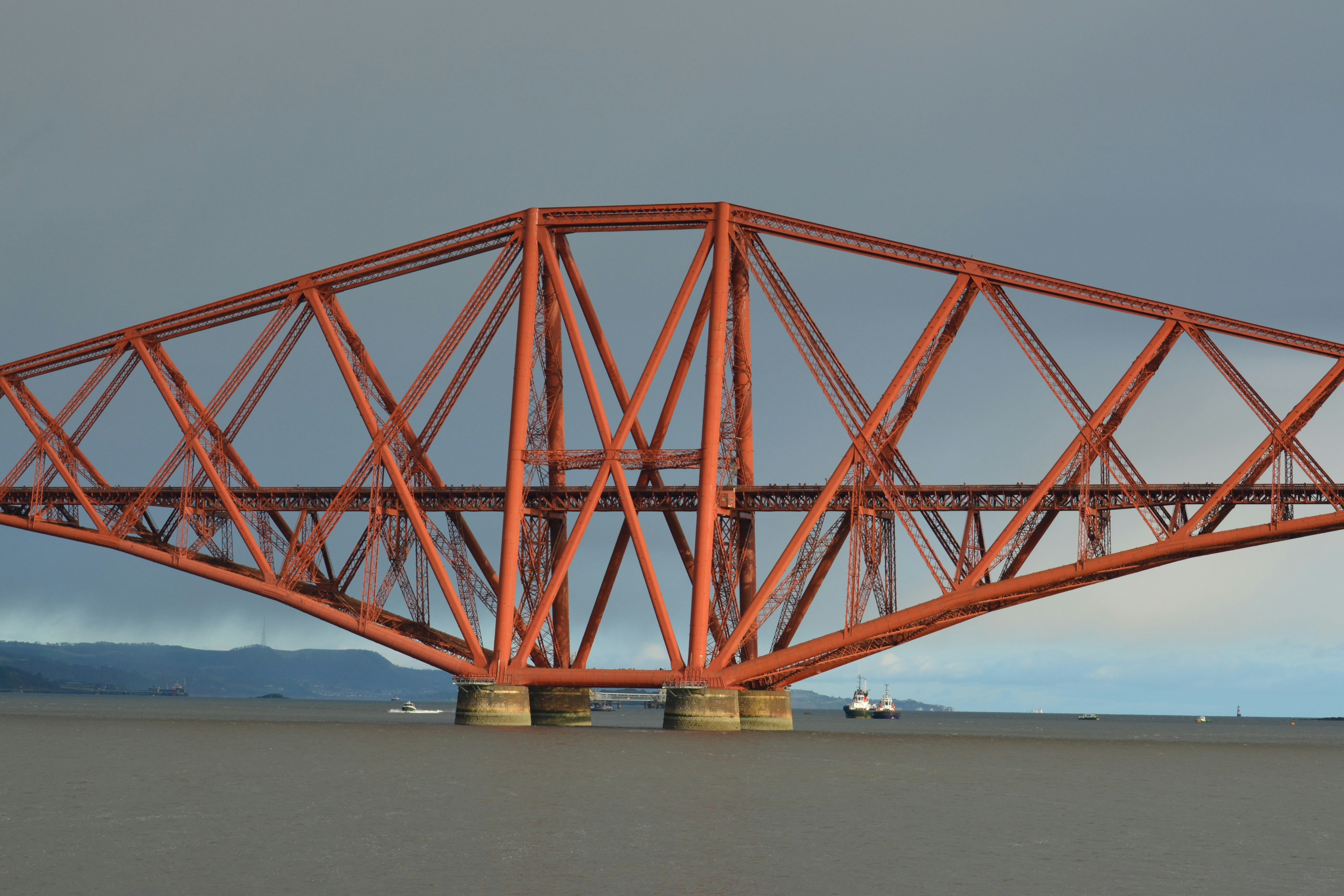 A striking close-up view of a massive cantilever tower of the iconic Forth Bridge spanning the Firth of Forth estuary in Scotland. The image highlights the intricate red steel lattice framework and industrial engineering of this famous UNESCO World Heritage Site. In the background, small boats dot the grey-brown water beneath a heavy, moody overcast sky, while the distant shoreline sits along the horizon. This perspective perfectly captures the immense scale, structural geometric complexity, and historic brilliance of Scotland's legendary railway bridge.