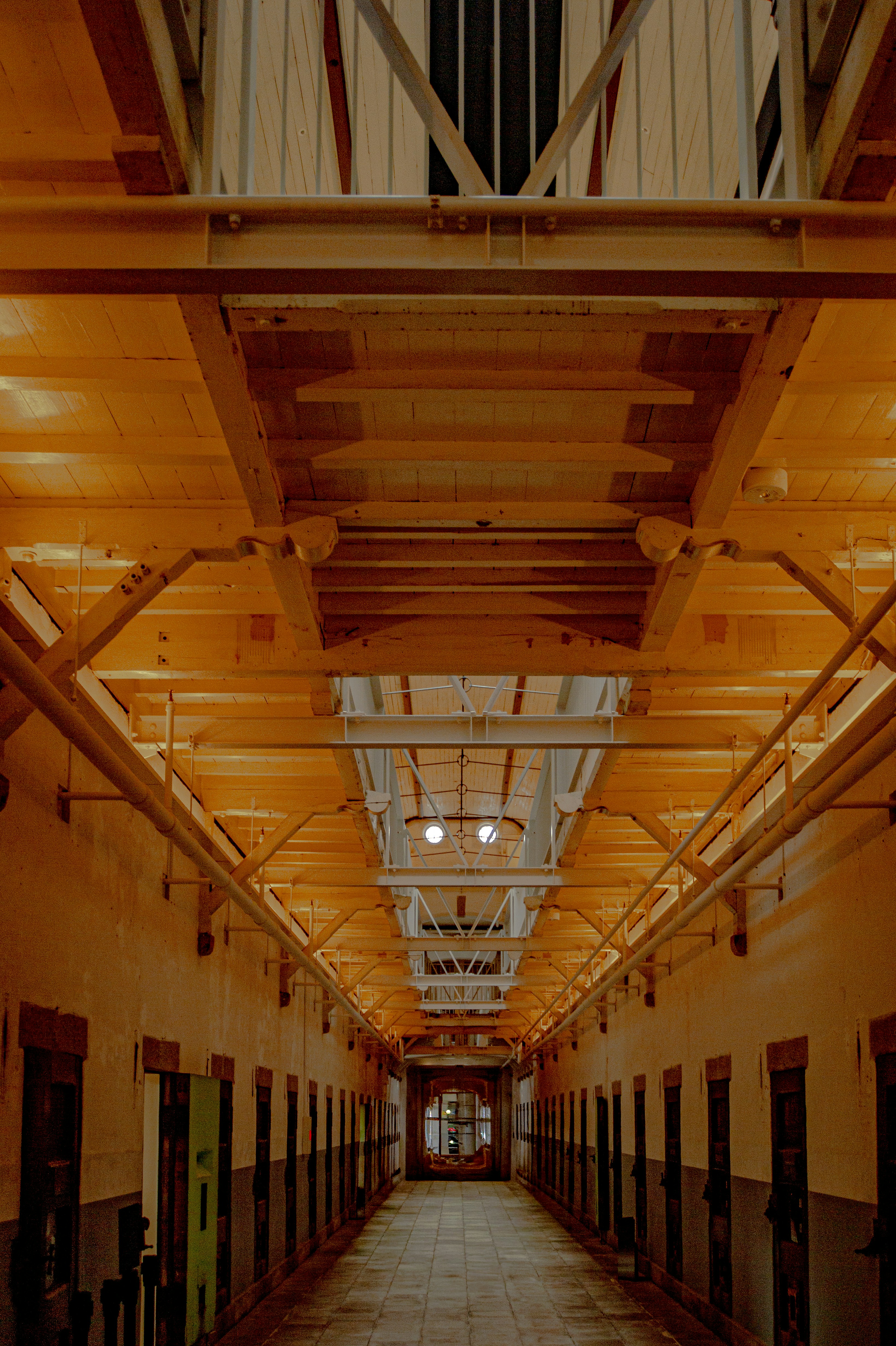 A long, dim corridor inside a historic building, with warm amber tones and repeating doorways along both sides, drawing the viewer’s eye toward a distant arched opening. The scene emphasizes architecture and light in an industrial ruin.