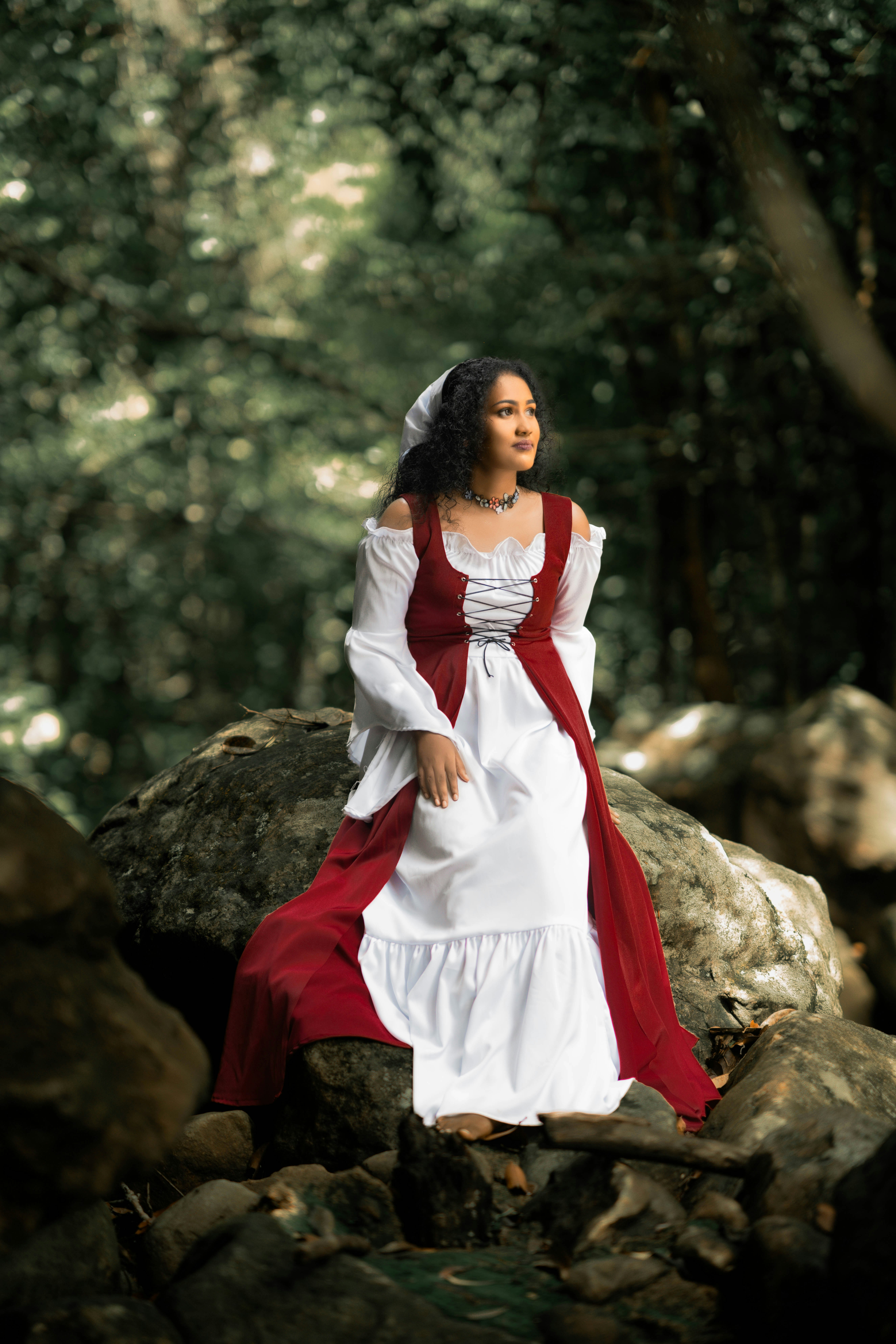 Woman in period dress sits on rocks in a forest.