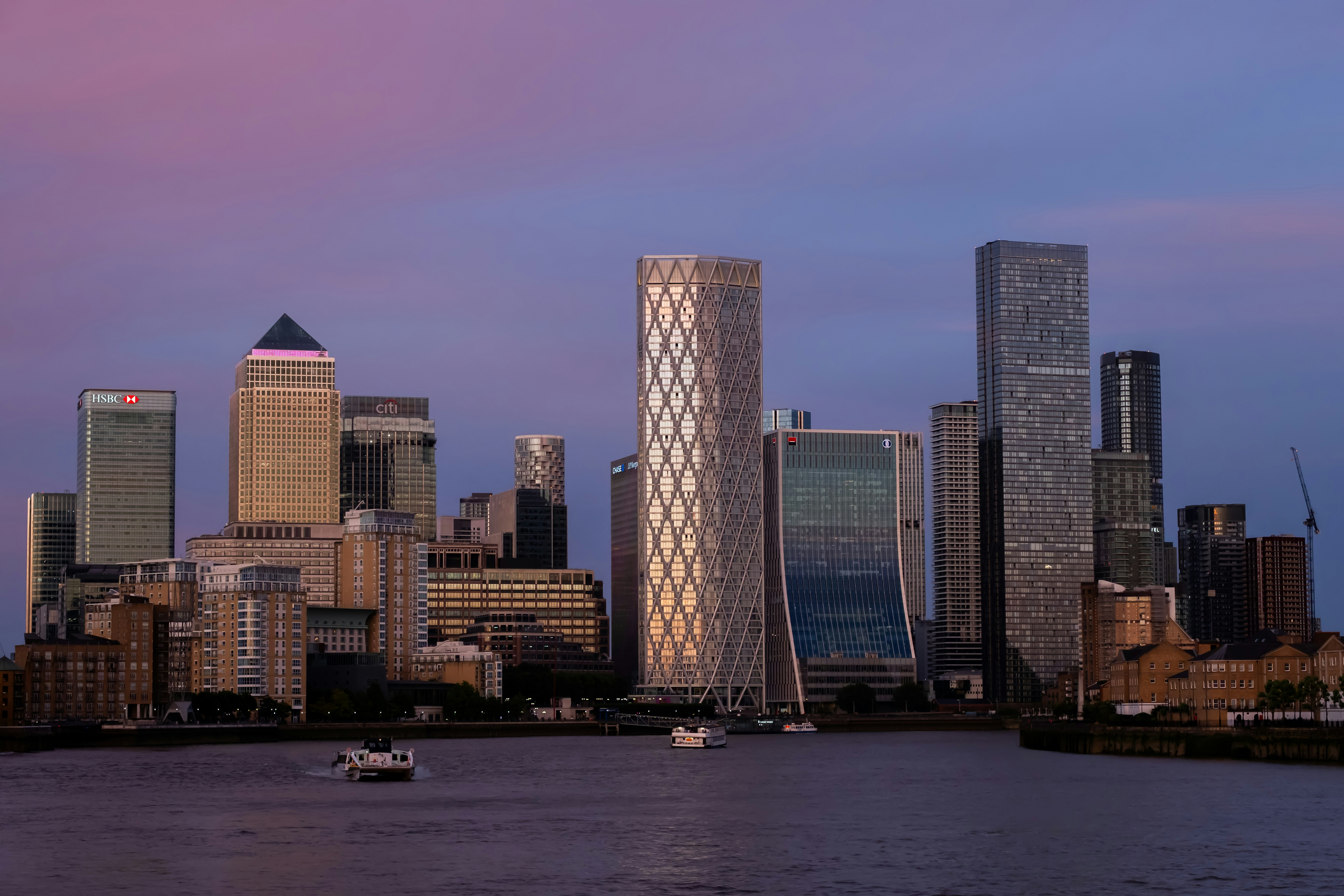 Modern cityscape at sunset with river and boats.