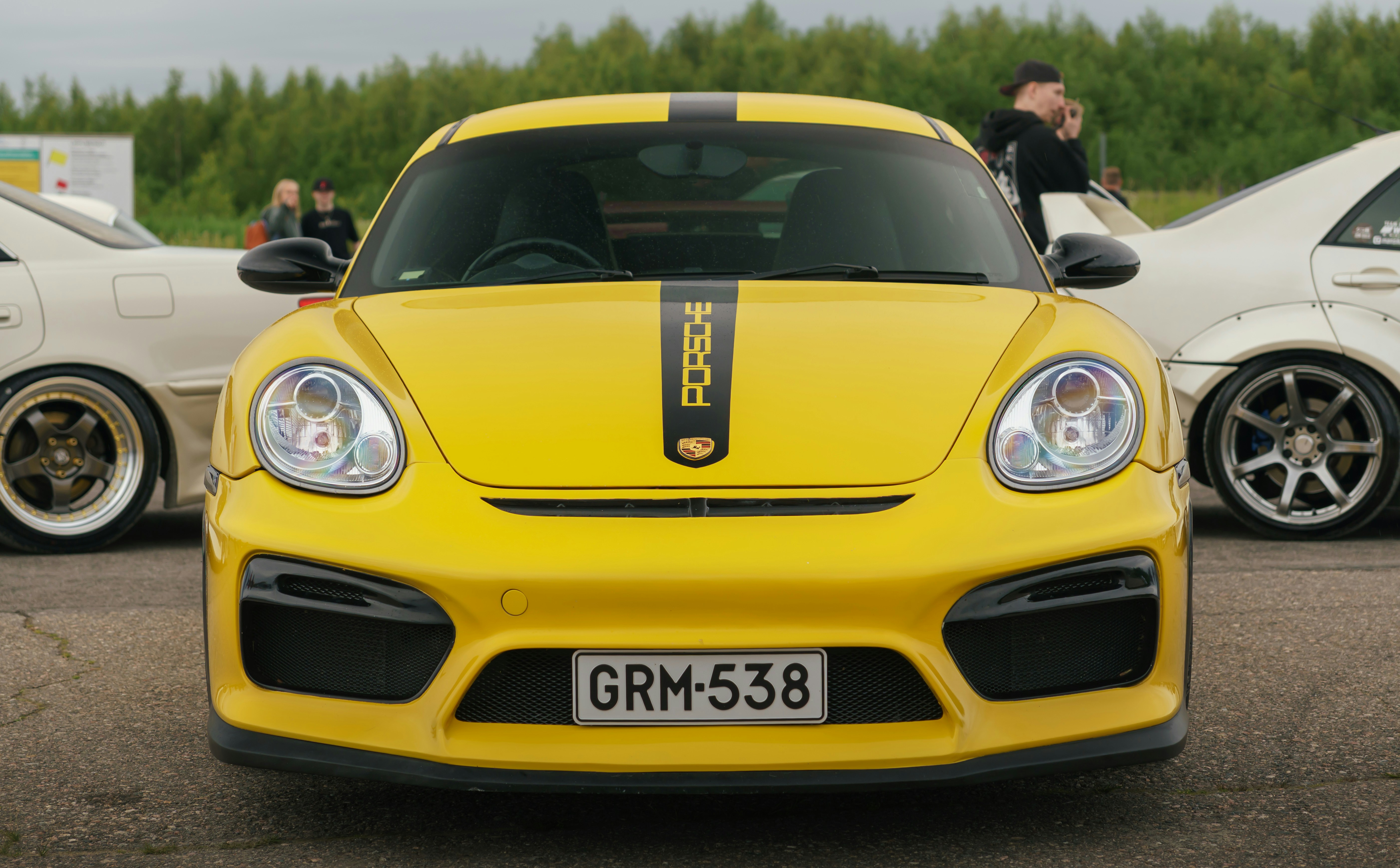 Bright yellow Porsche at a car meet, frontal view with license plate and people in the background; a realistic automotive shot highlighting the front fascia and stance.