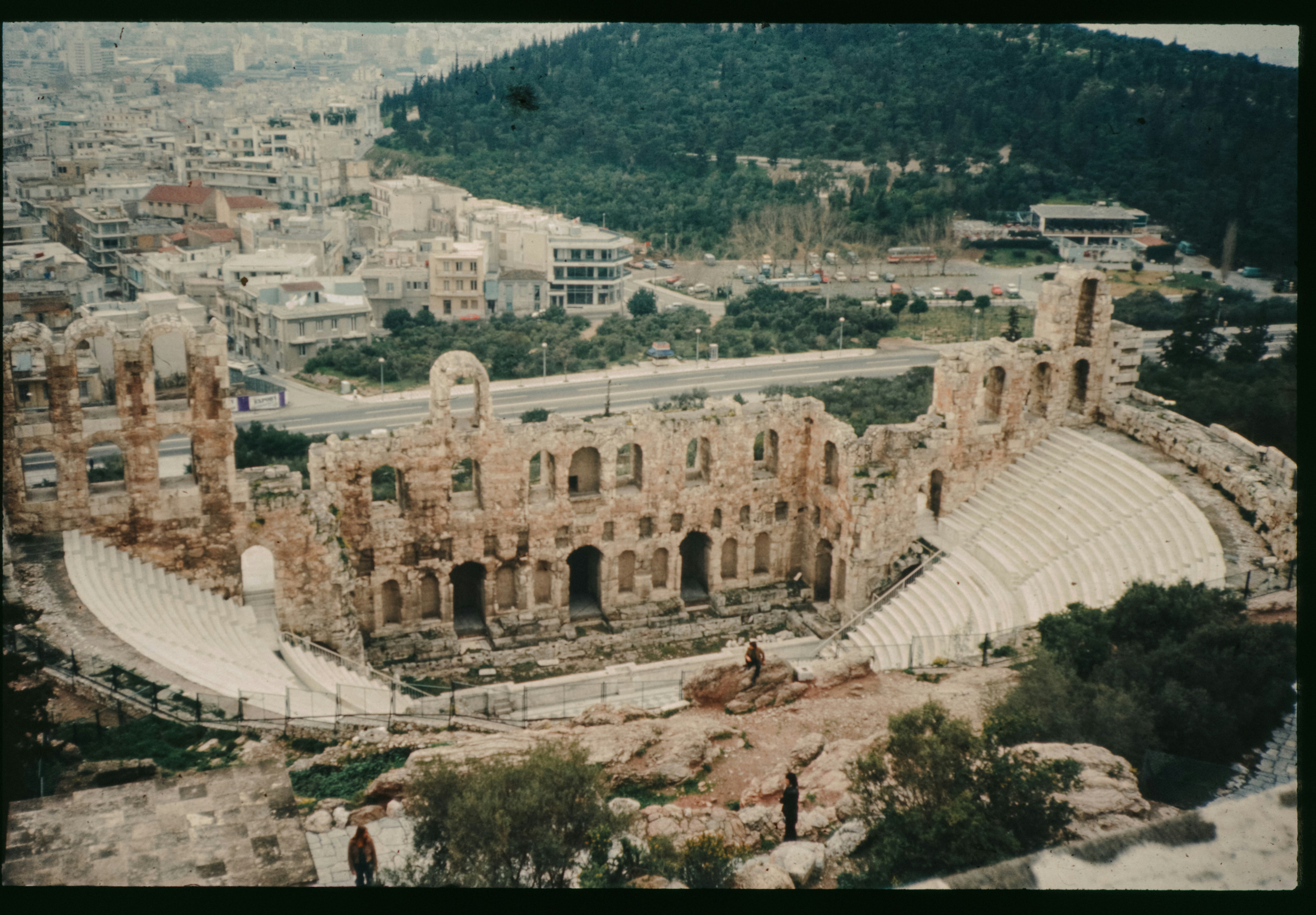 Ancient stone theater ruins with a city and forested hill.