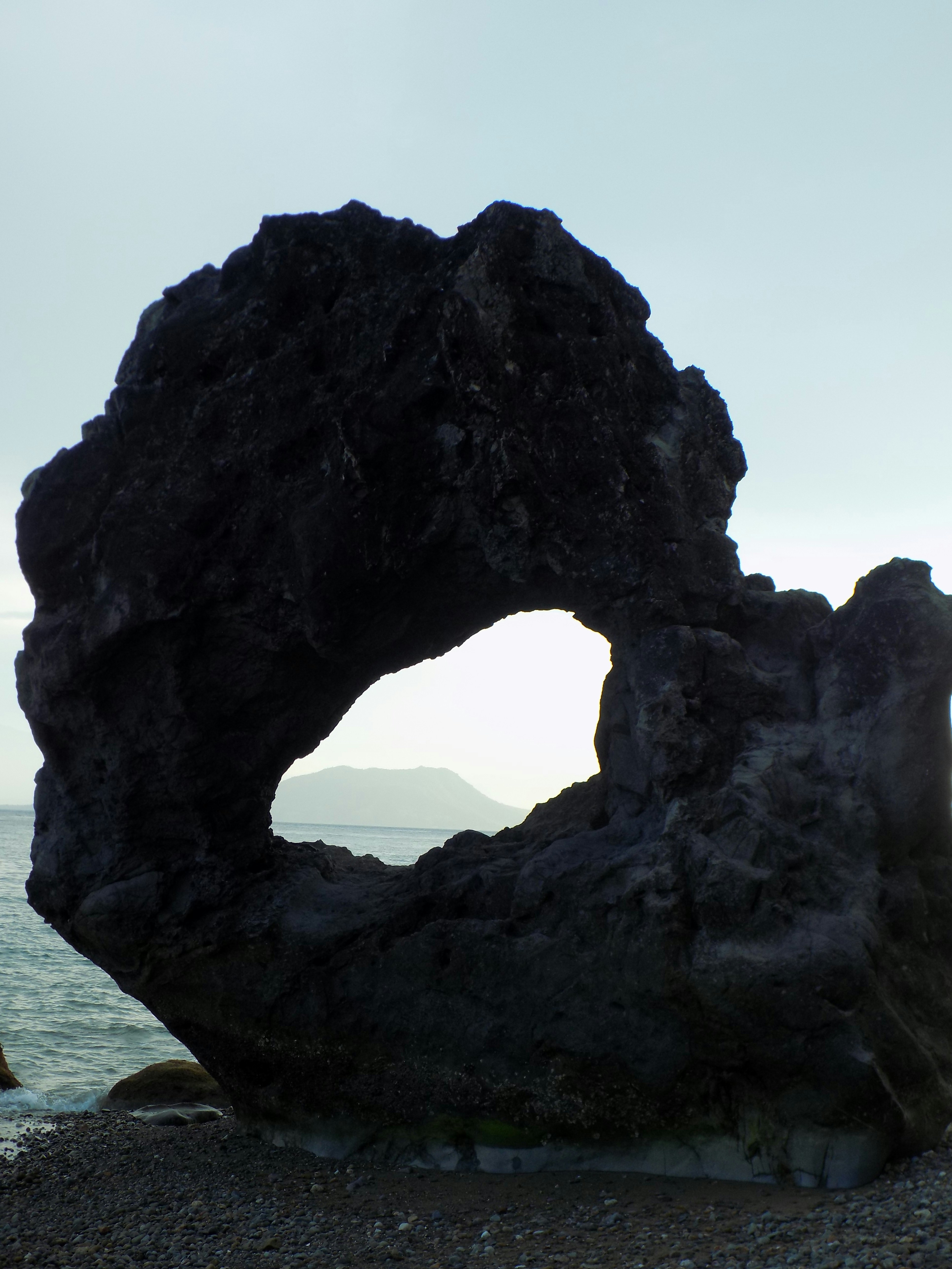 Rugged rock arch by the shoreline with a luminous gap revealing distant land; a minimalist seascape emphasizing texture and negative space.