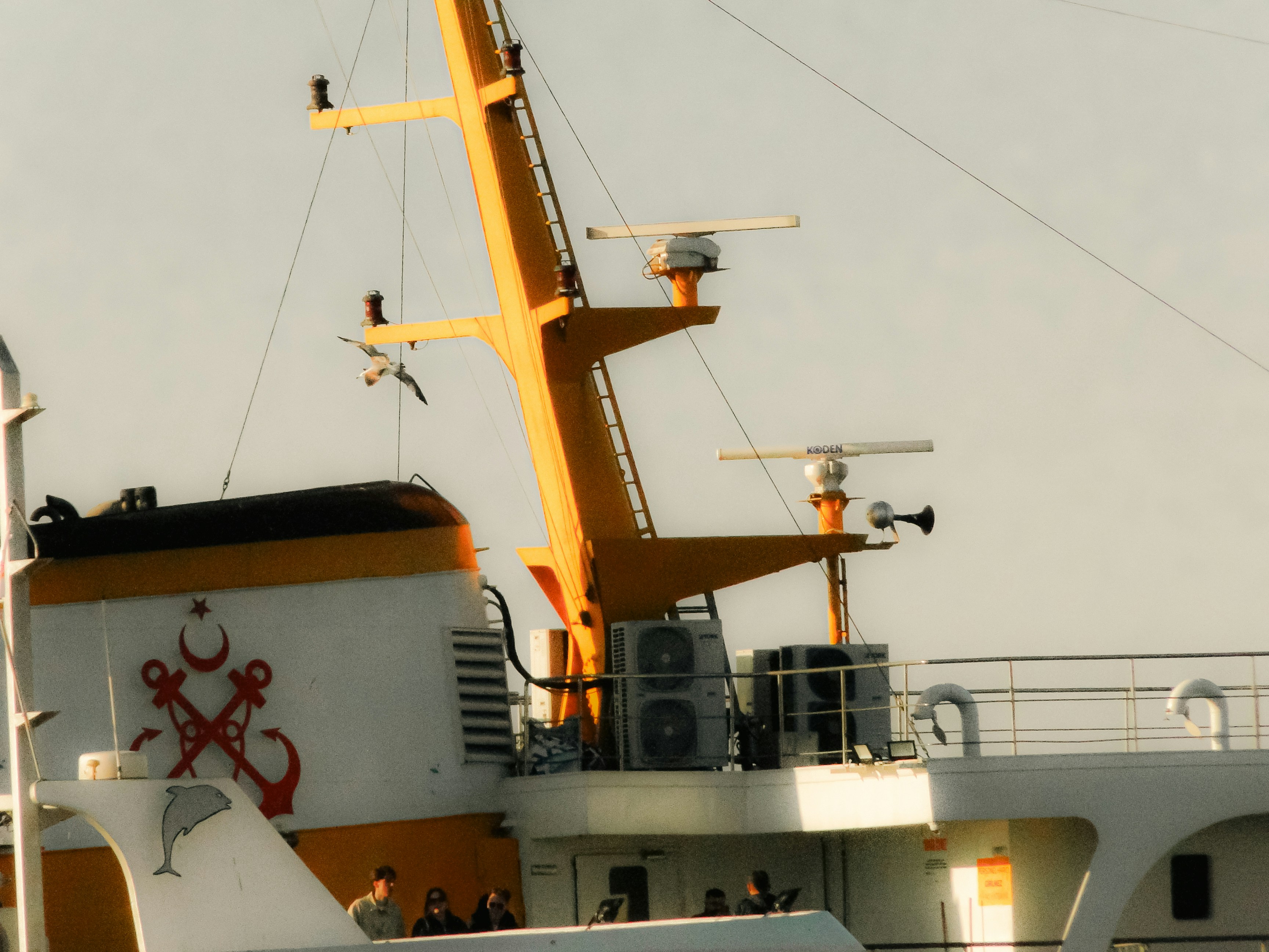 Crew deck of a ship with orange-yellow superstructure and railings against a pale sky; silhouettes of people near the lower deck and visible equipment on the upper deck.