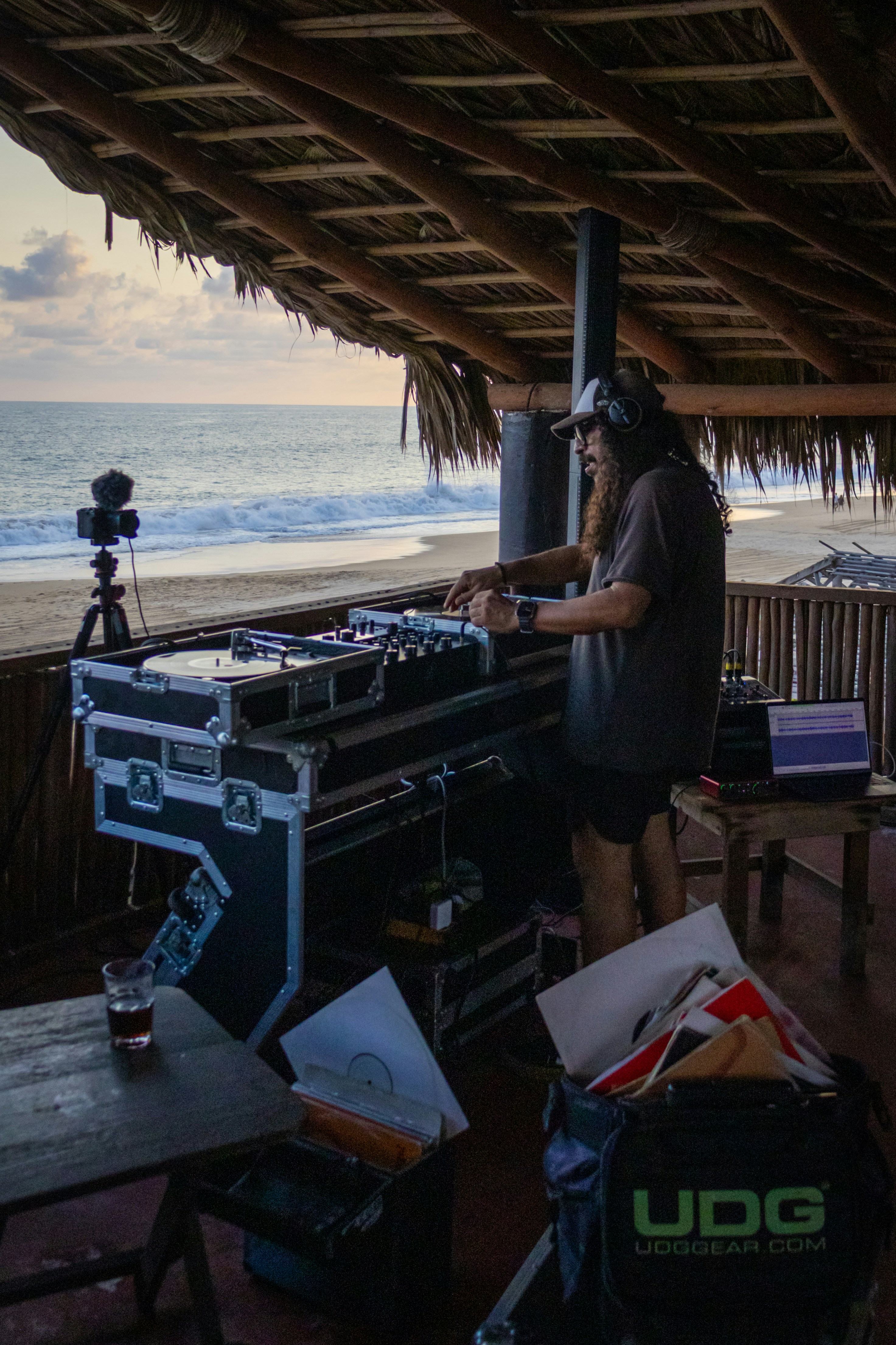 DJ setup on a seaside deck with waves in the background and a person adjusting equipment under a thatched roof.