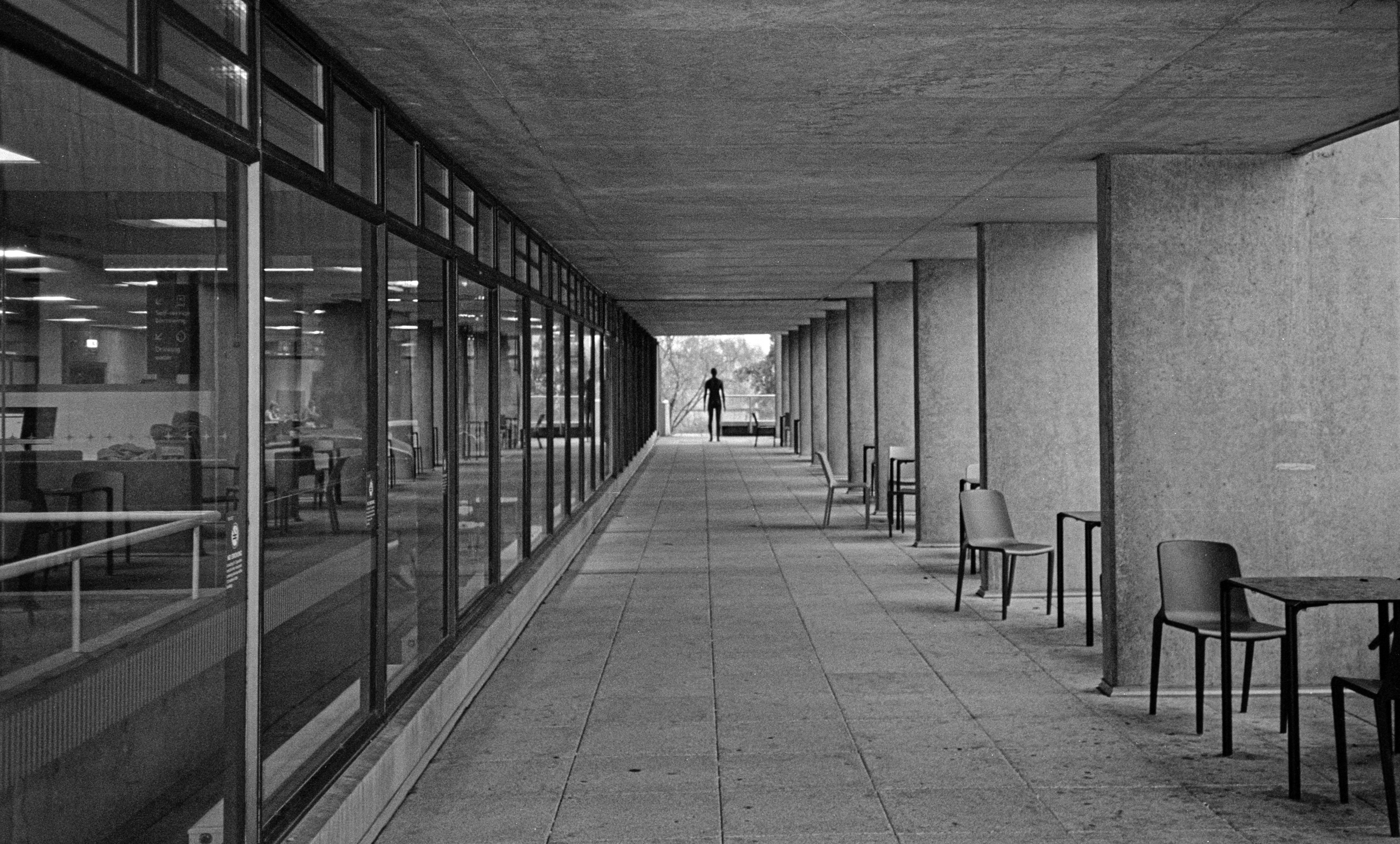 A lone figure stands at the end of a long brutalist concrete colonnade in black and white.