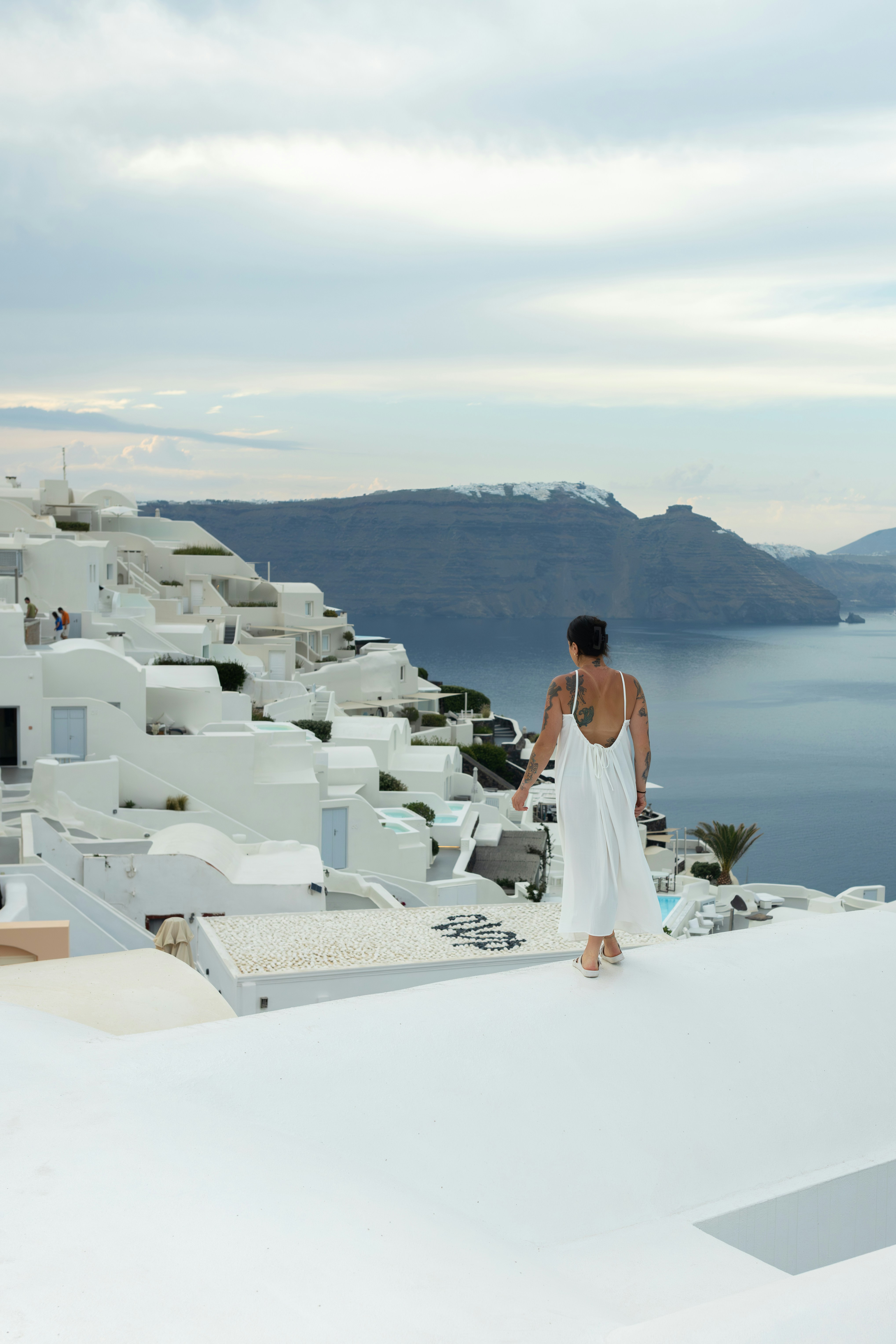 A lone figure in a white dress gazes toward the Aegean from a sunlit white-washed terrace