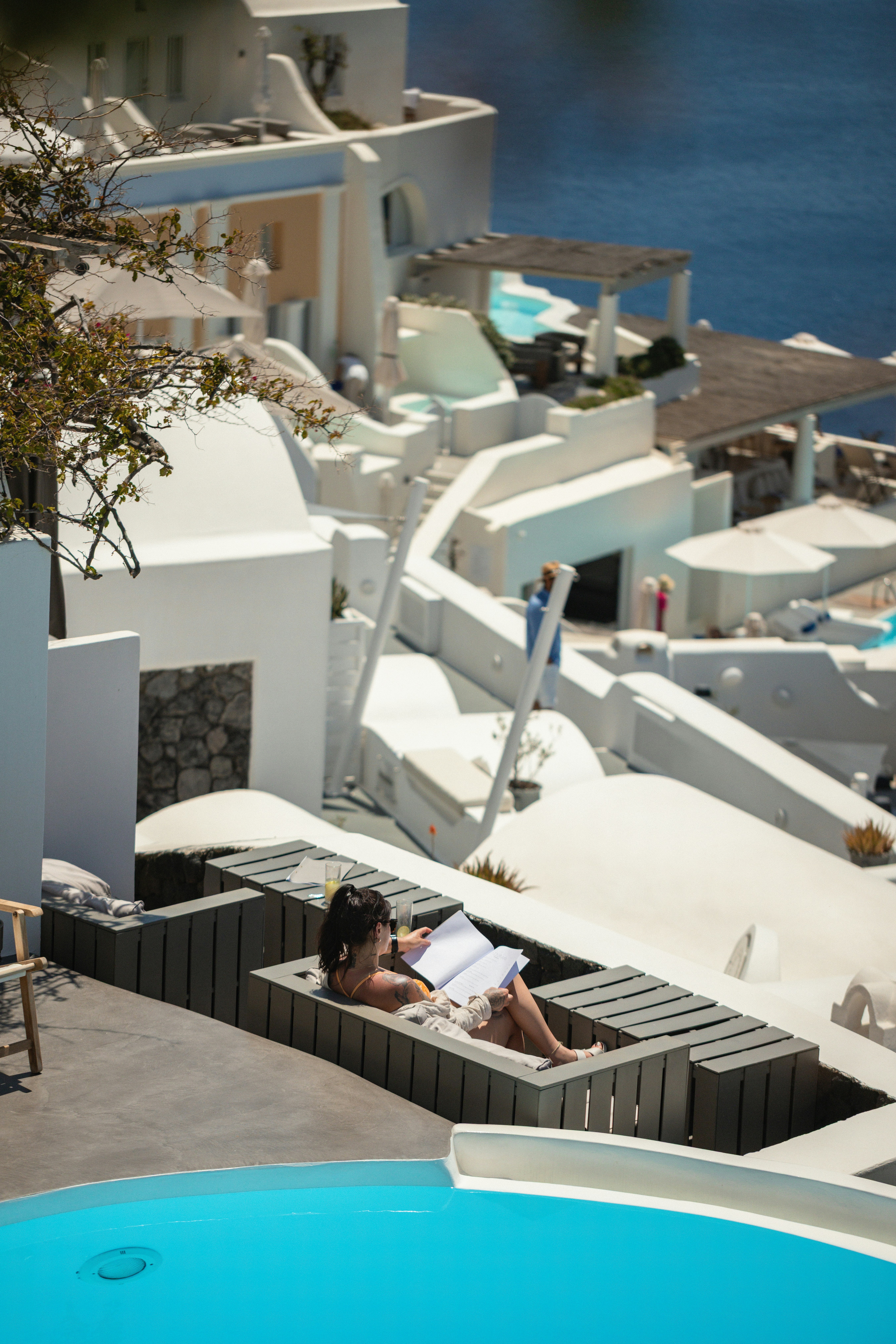 A sunlit rooftop lounge overlooks a blue pool, where a person reclines on modular seating reading a book, with whitewashed terraces and sea views in the background.