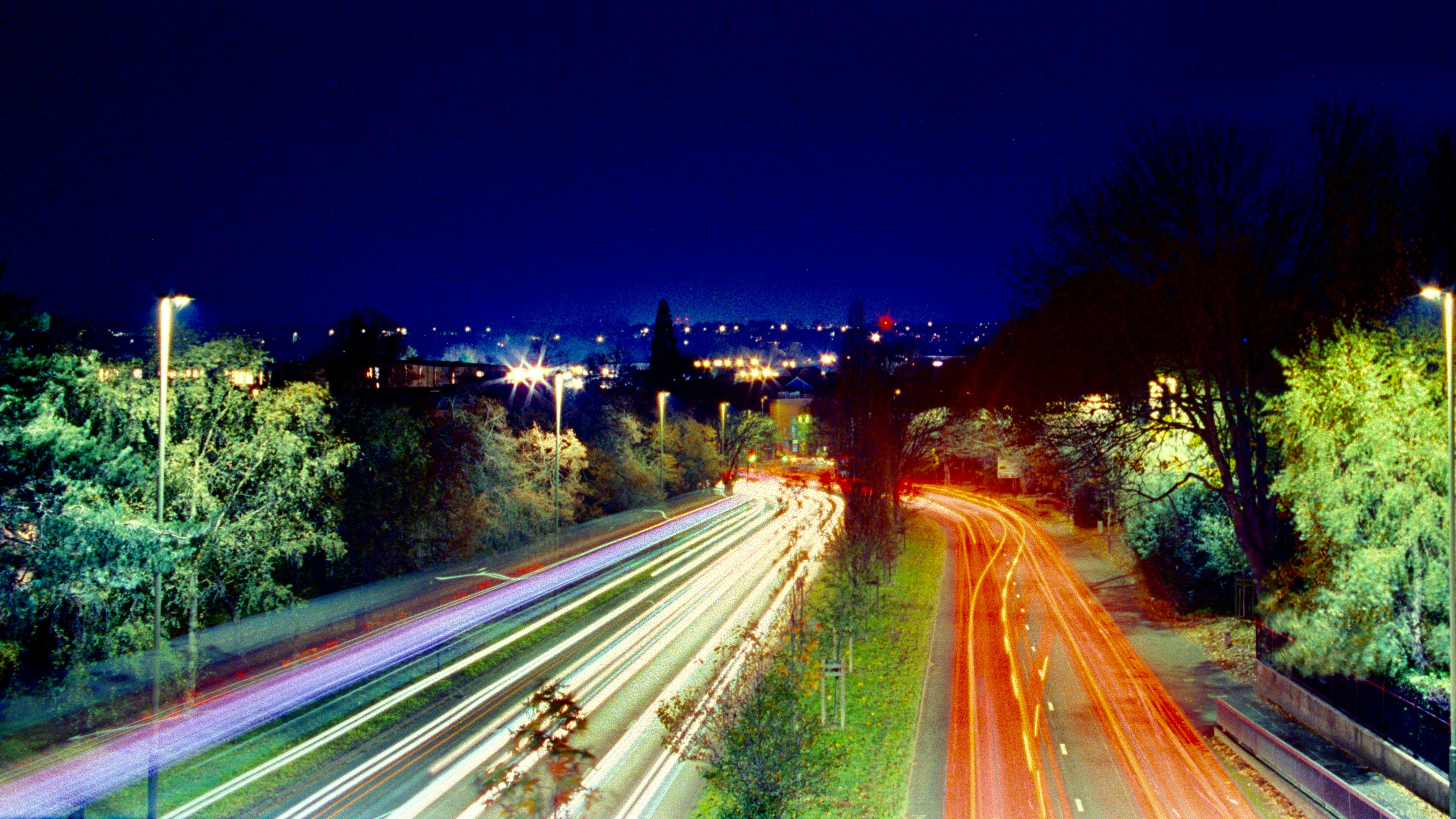 Long-exposure light trails streak along a tree-lined dual carriageway at night.