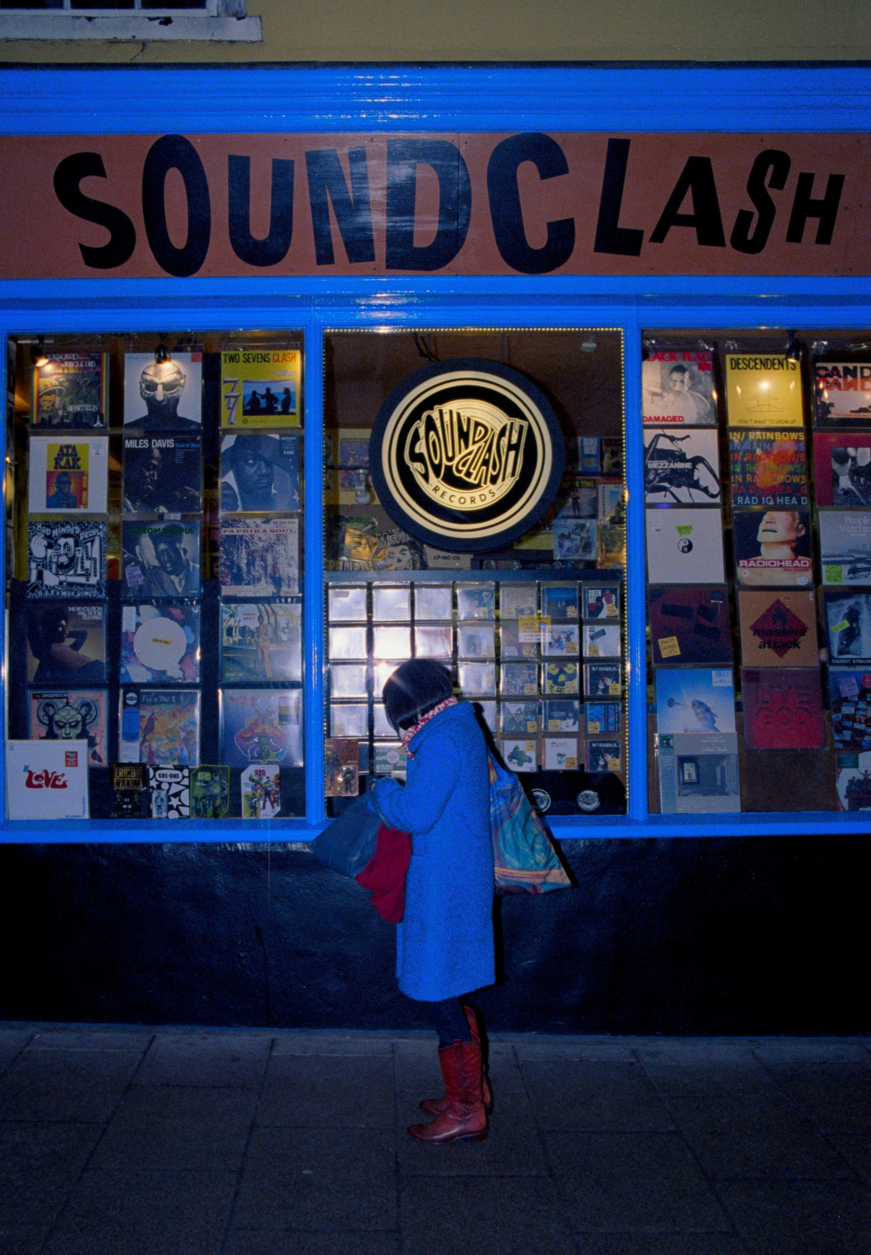 A person in a blue coat browses the neon-lit window of a record shop at night.