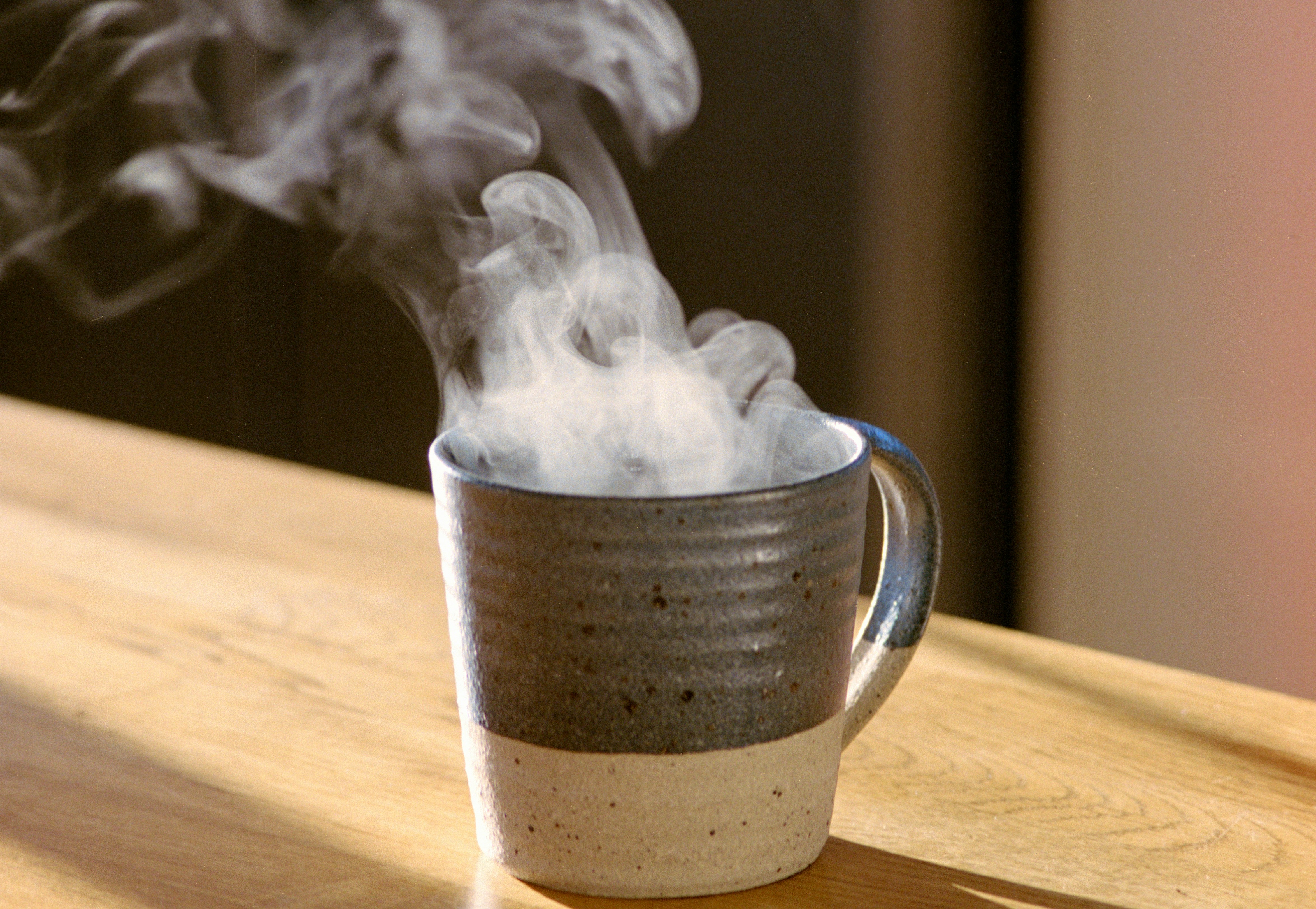 Steam rises from a two-tone ceramic mug on a sunlit wooden table.