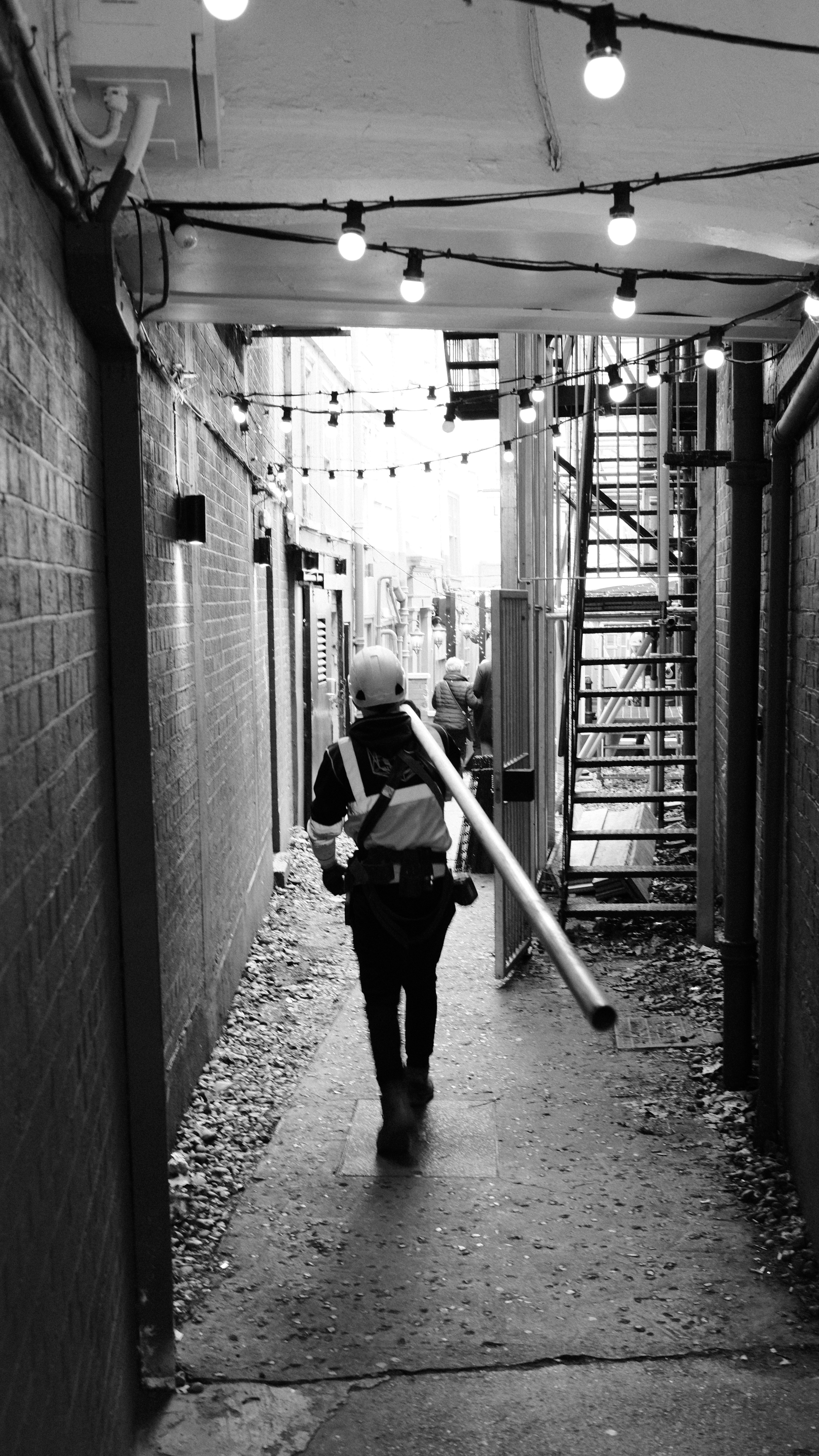 A worker shoulders a long pole down a narrow alley strung with festoon lights.