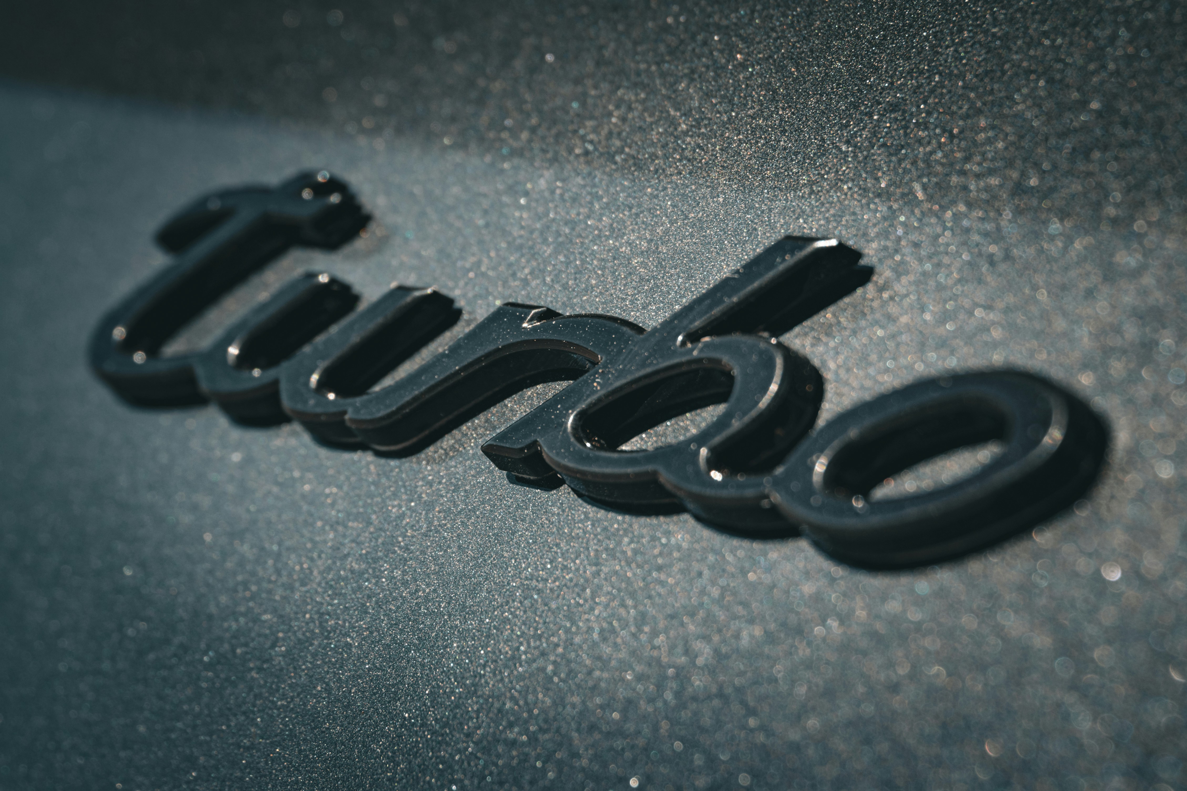 A glossy black "turbo" script emblem on metallic flake paintwork, sparkling under soft light. Macro detail celebrating forced-induction badging.