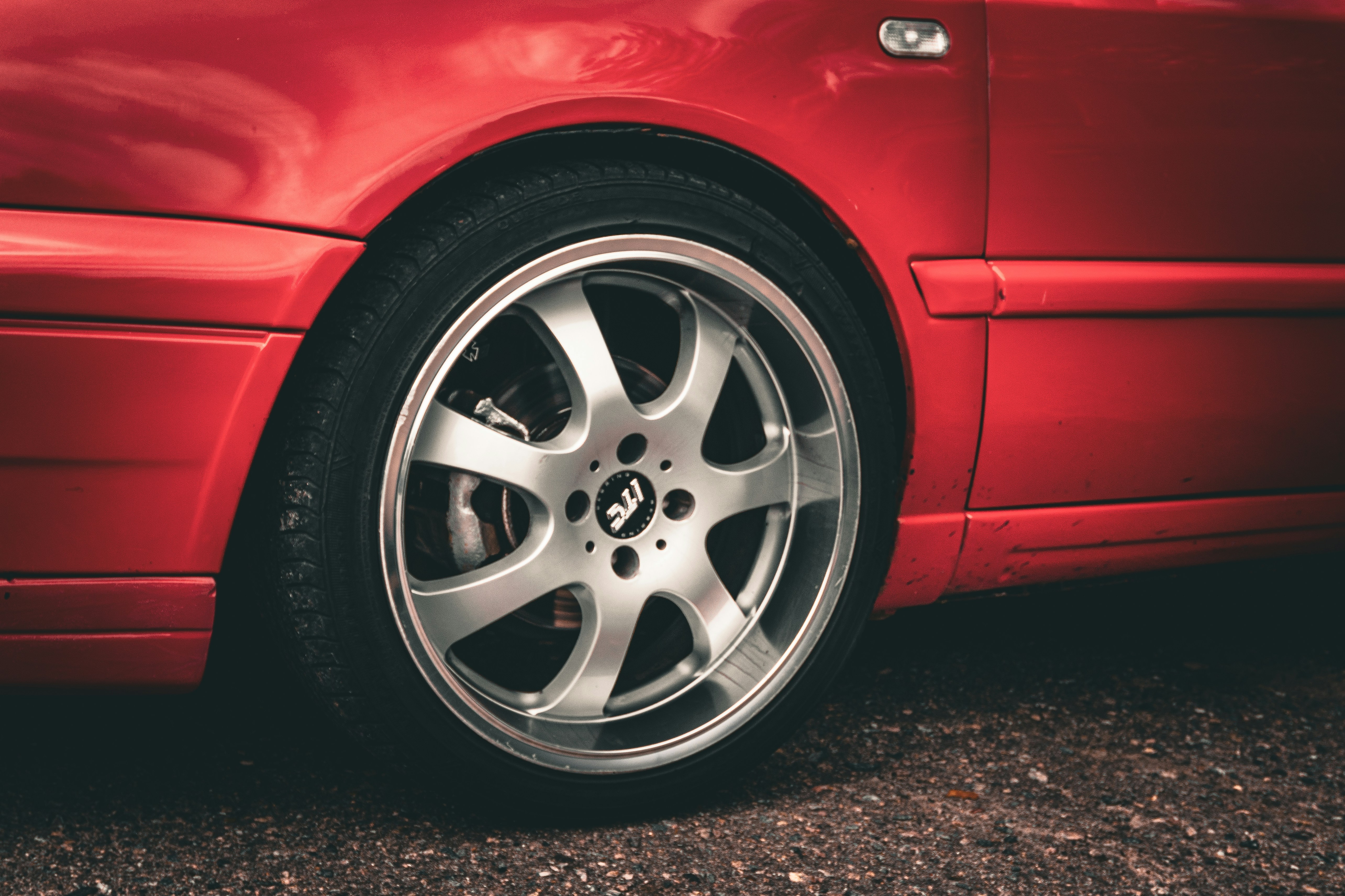A polished seven-spoke alloy wheel on a glossy red sedan, low-profile tire hugging the arch. A clean side-on detail of a lowered car on tarmac.