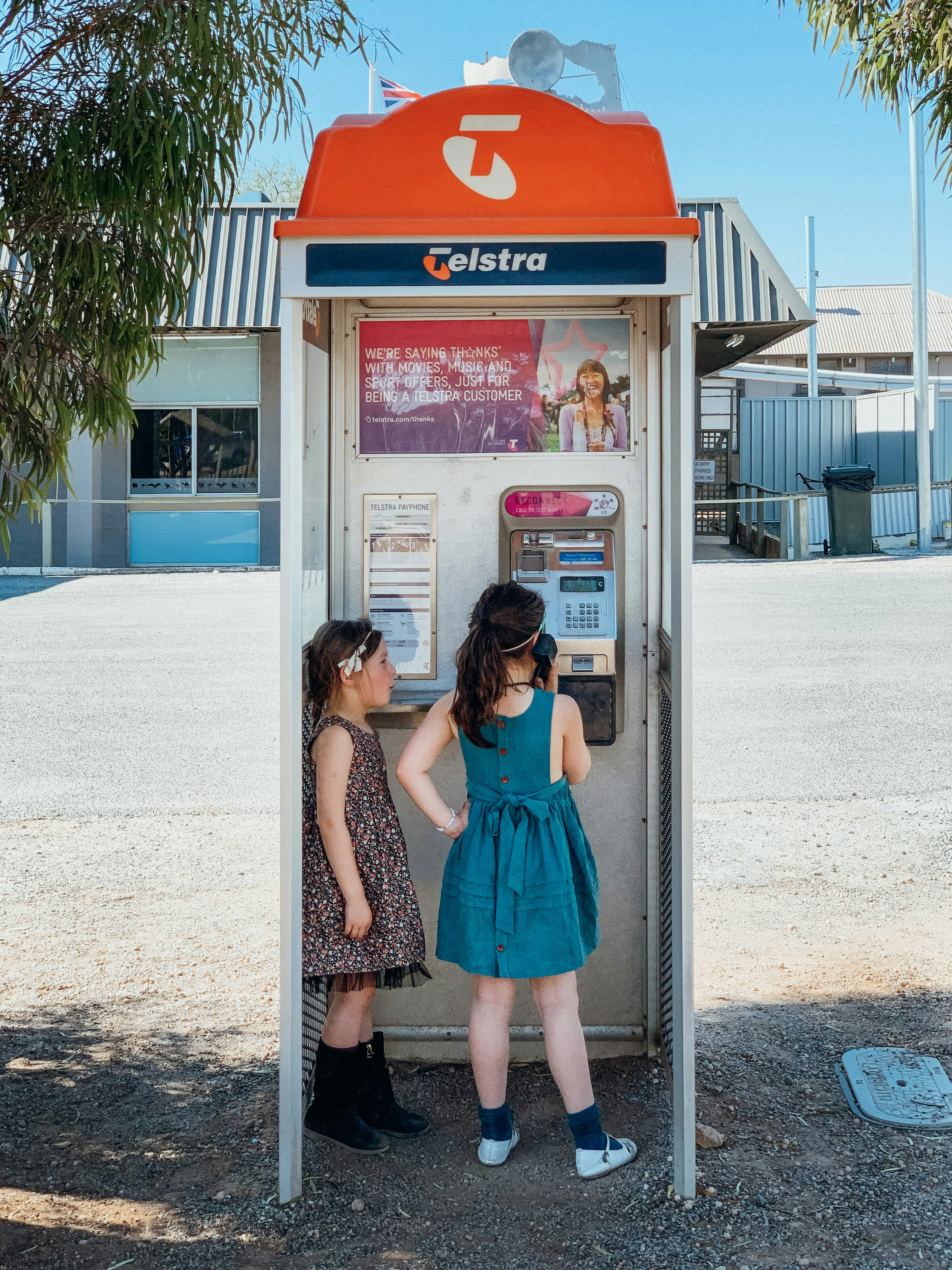 Children at a Telstra kiosk: sunlit curiosity under a bright orange roof