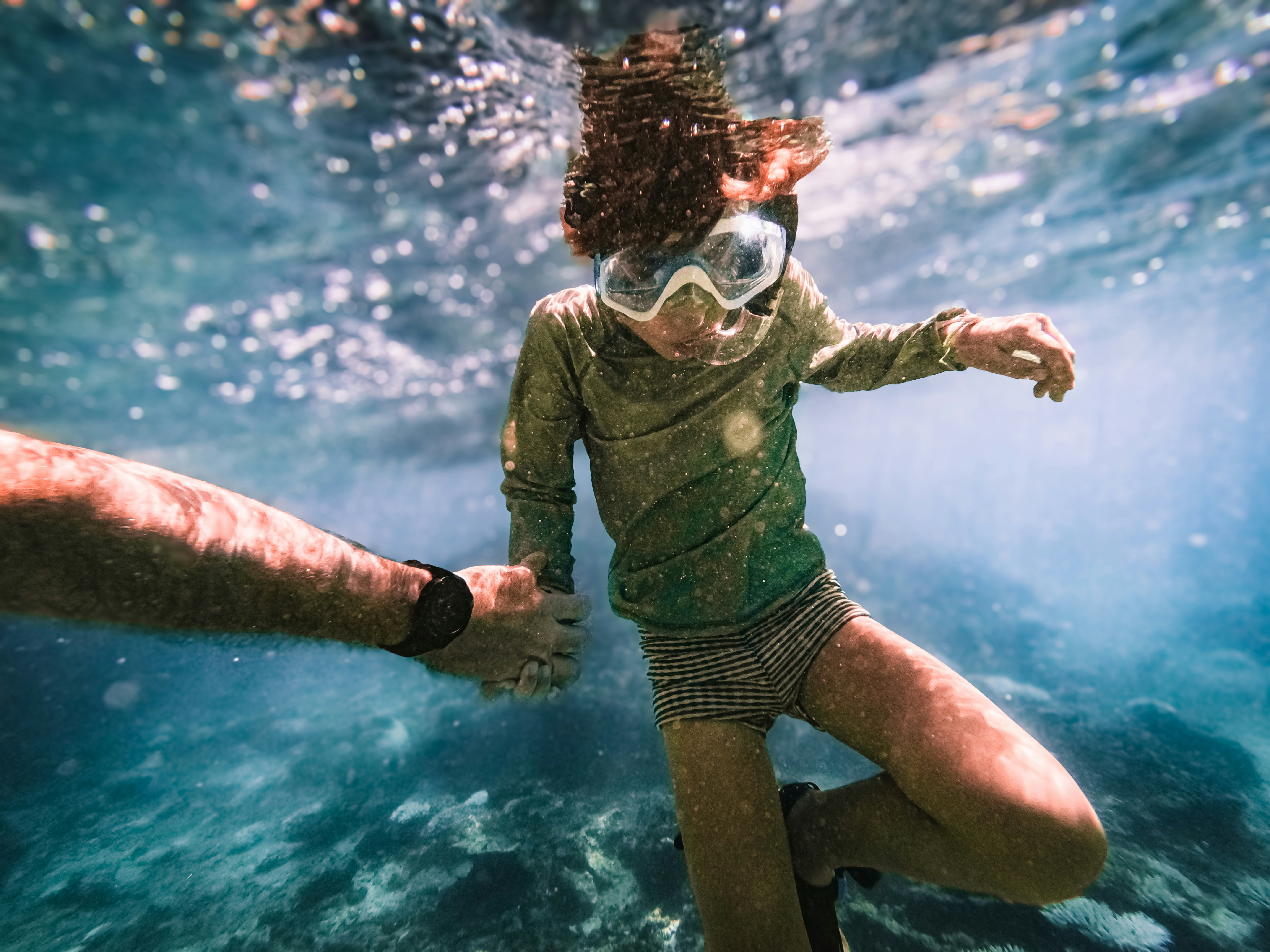 Young snorkeler reaches for a guiding hand in clear blue water, wearing snorkel mask and striped swim shorts. An arm of another diver is visible, creating a sense of connection. The scene emphasizes underwater exploration and trust.