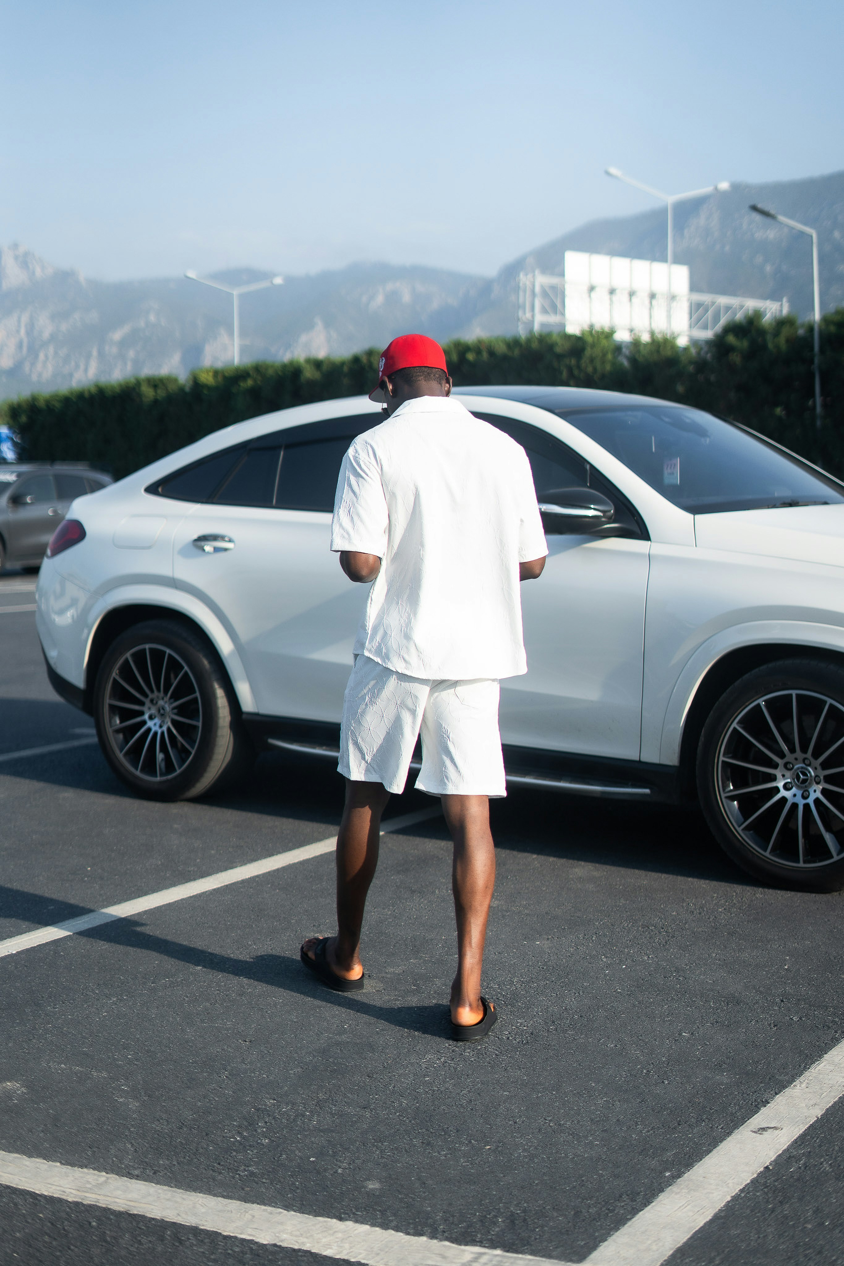 Man in white outfit stands beside a white SUV in a sunlit parking lot