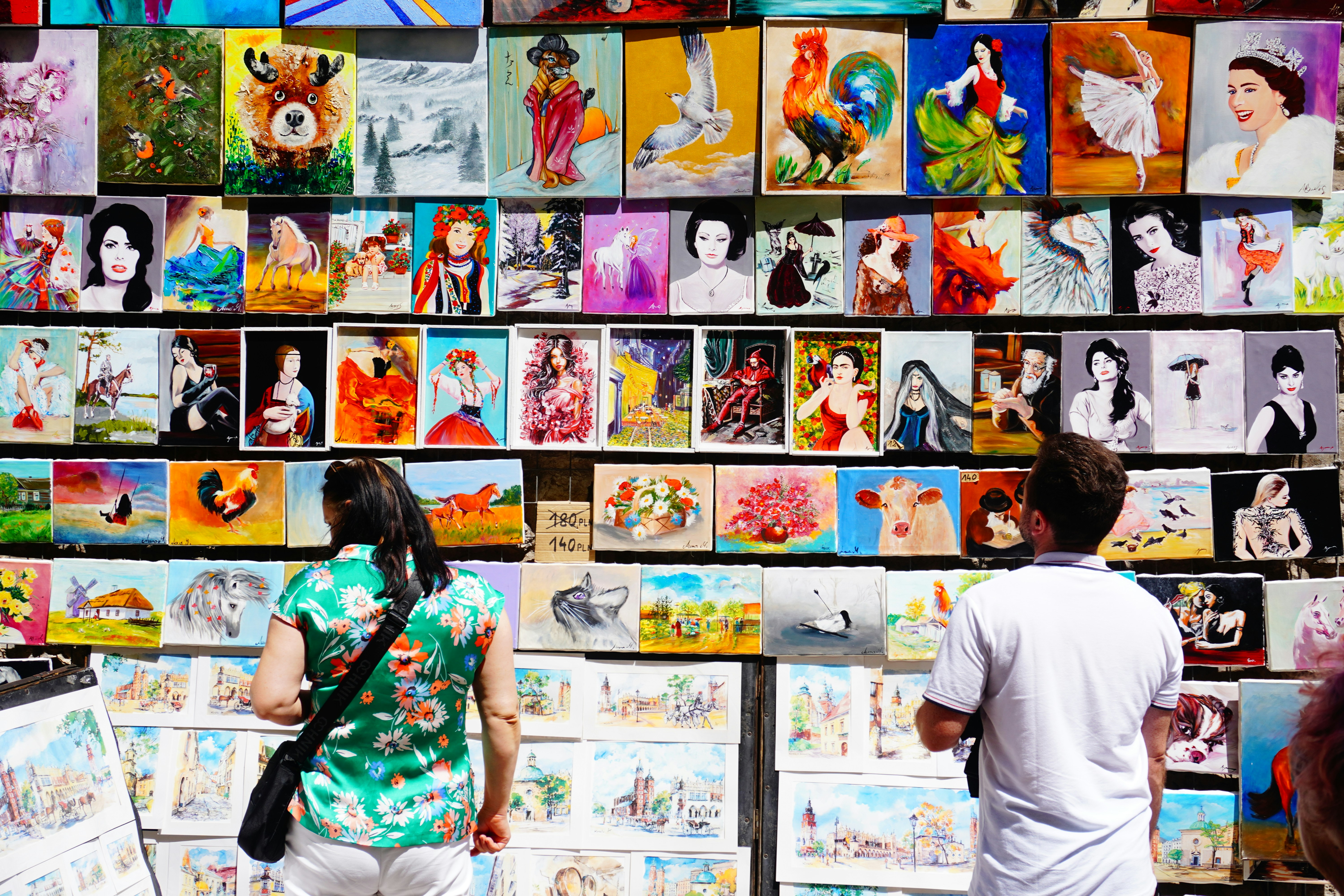 People viewing many colorful paintings displayed on a wall.