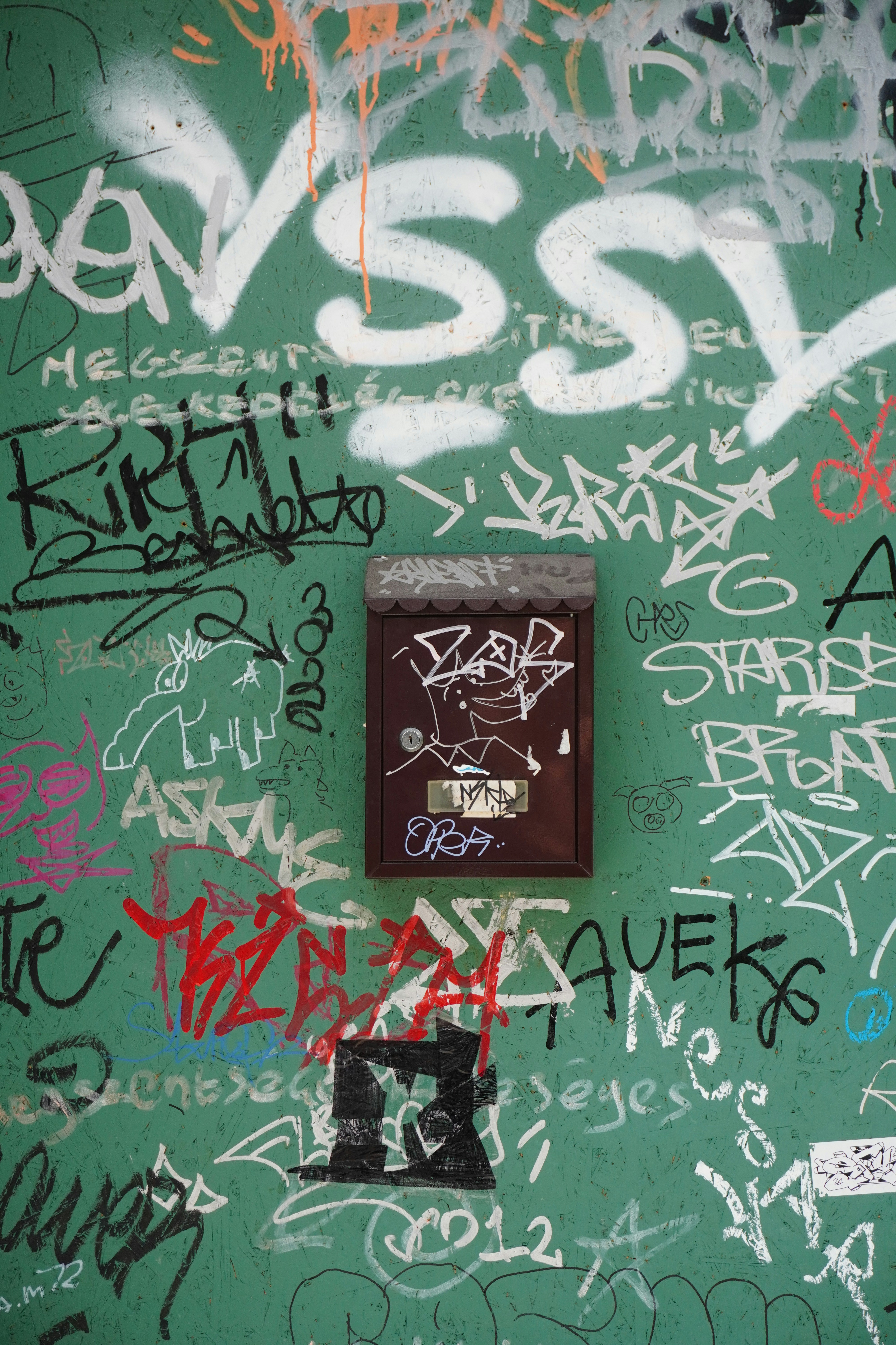A brown mailbox on a green wall covered in graffiti.