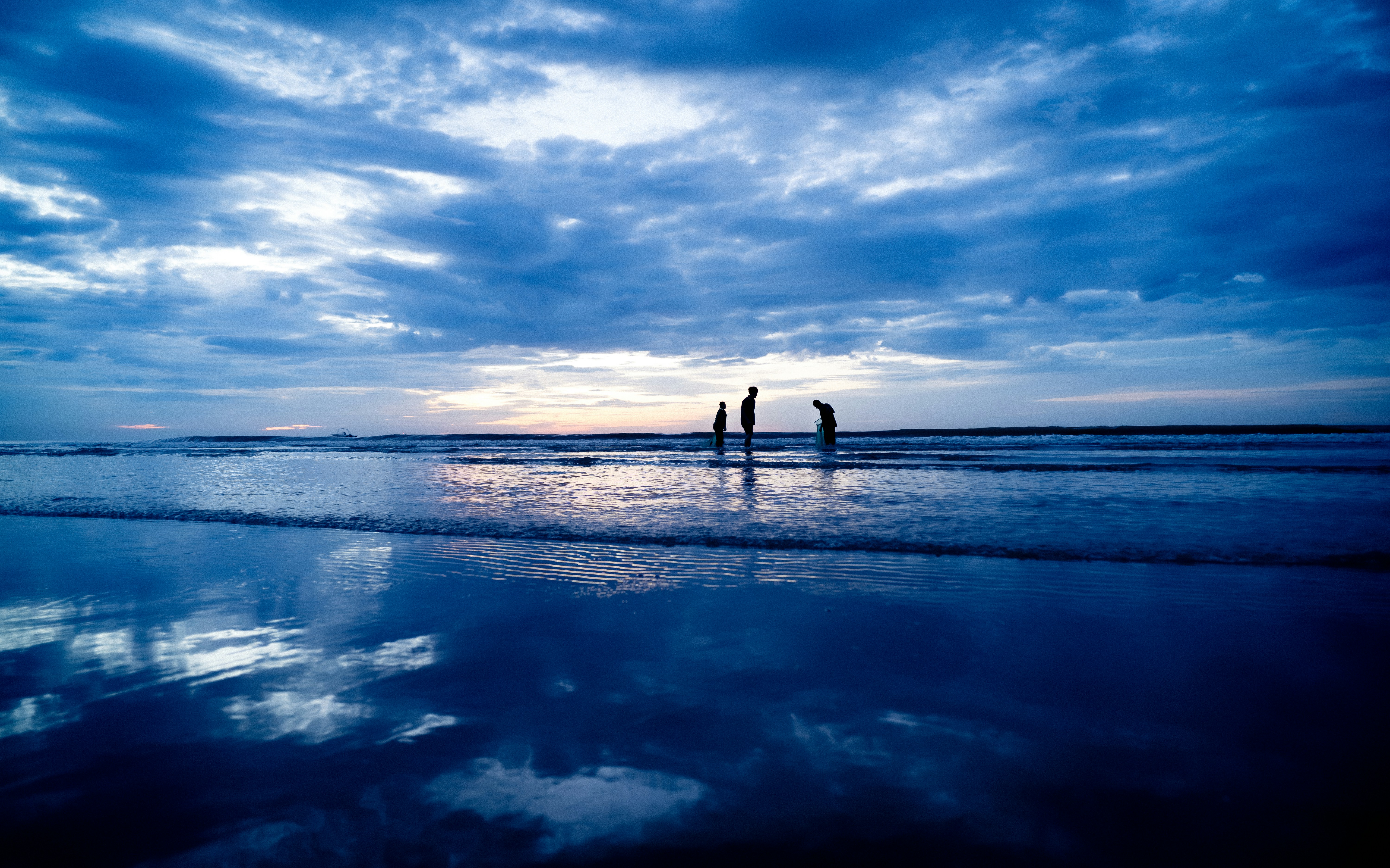 A breathtaking, cinematic shot of three silhouetted figures standing in the shallow waters of a vast beach under a heavy, dramatic sky. The scene is dominated by striking deep blue tones, creating a serene yet melancholic atmosphere. The calm tide perfectly mirrors the moody clouds above, casting beautiful reflections onto the wet sand. In the far distance, a subtle hint of warm sunset light breaks through the horizon, offering a stunning contrast to the overwhelming blue palette. Perfect for themes of solitude, coastal beauty, and contemplative nature photography.