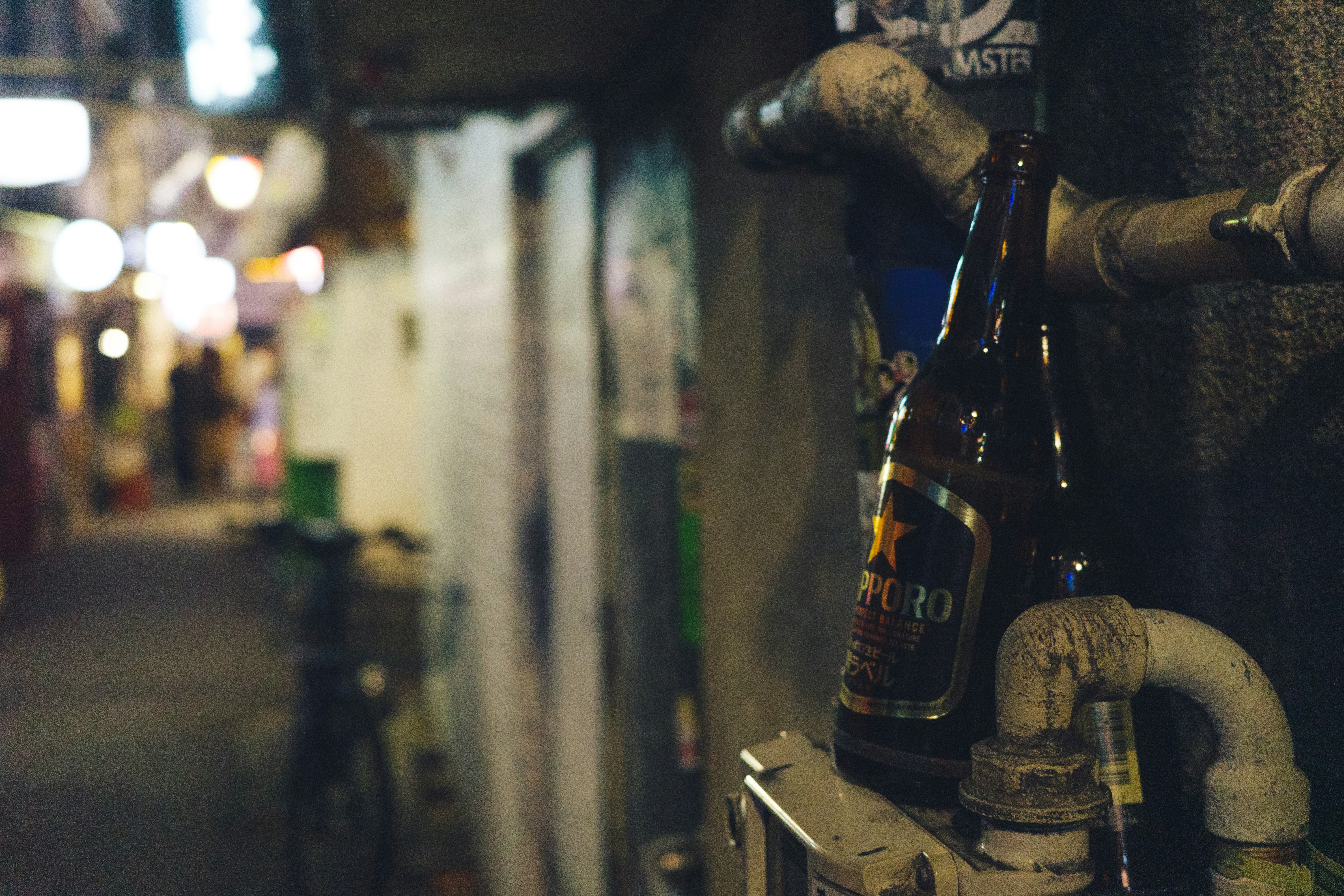A lone beer bottle sits atop a metal surface beside weathered pipes in a dim alleyway, with blurred background lights suggesting a distant cafe scene.
