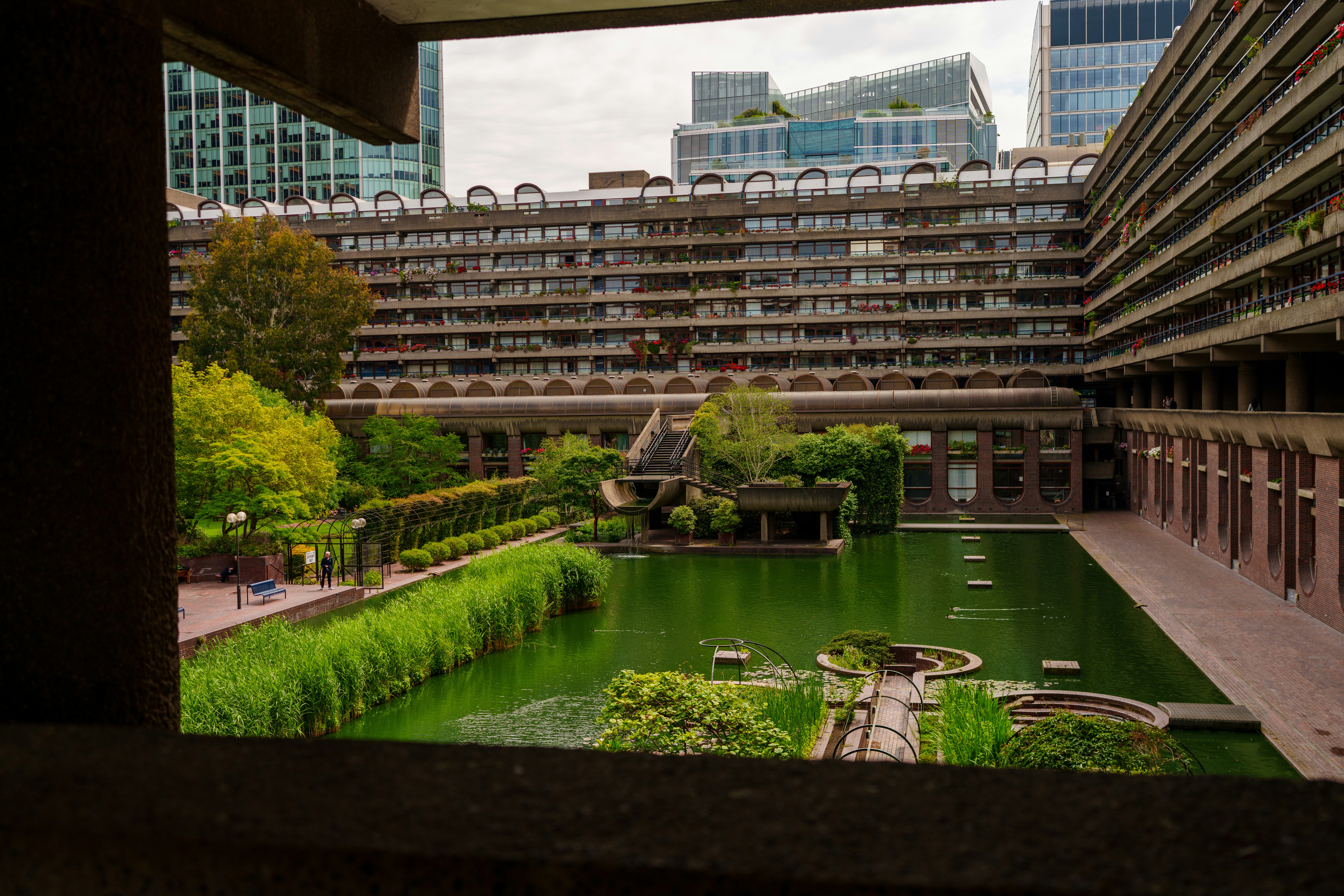 Brutalist architecture surrounds a vibrant green urban pond.