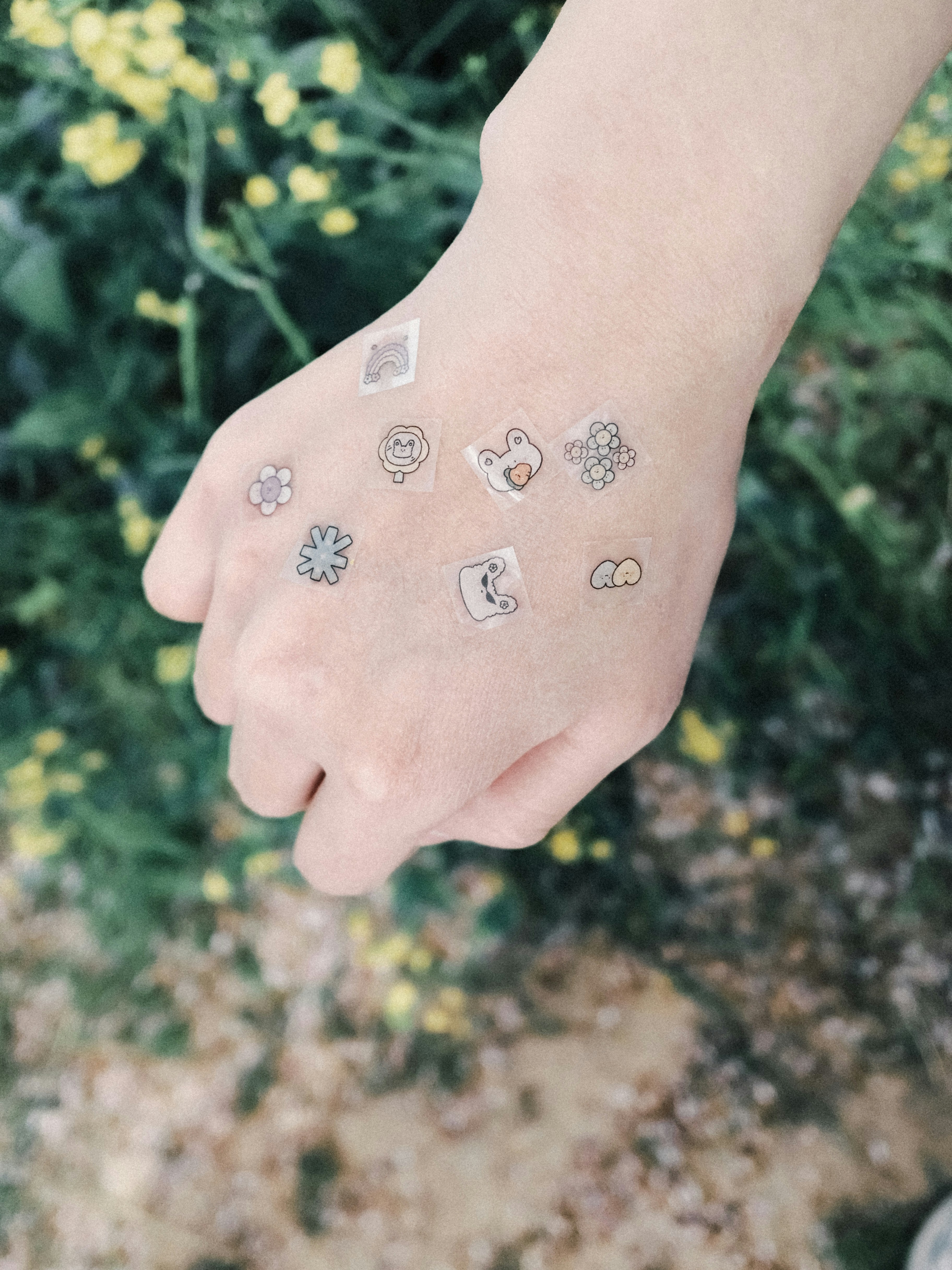 Close-up of a hand with small decorative stickers on the back, set against a blurred garden background featuring yellow flowers.
