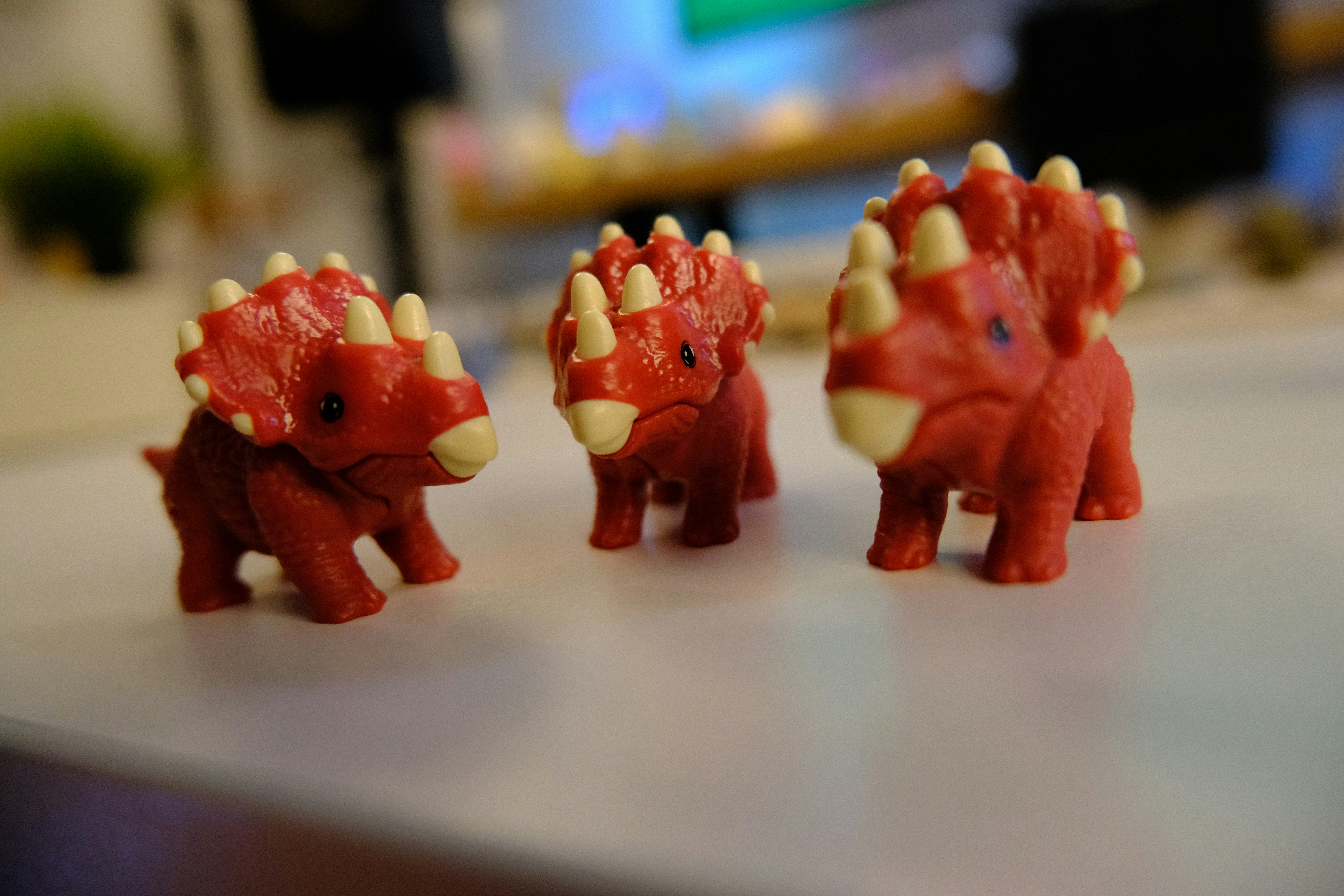 Three red toy triceratops figures stand on a white surface.