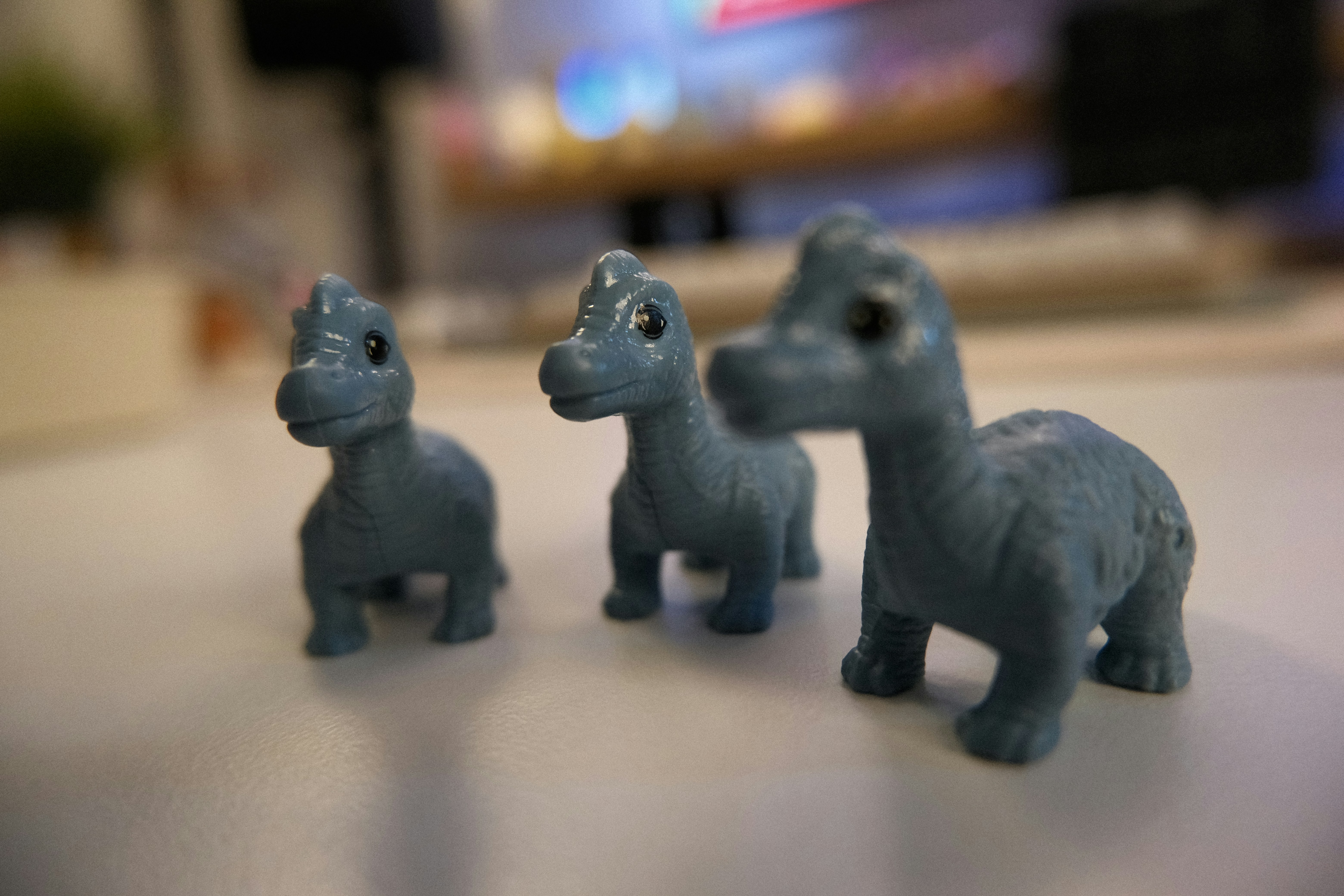 Three blue toy dinosaurs stand on a smooth surface with a blurred background, creating a shallow depth of field. The scene highlights small figures as the primary subject.