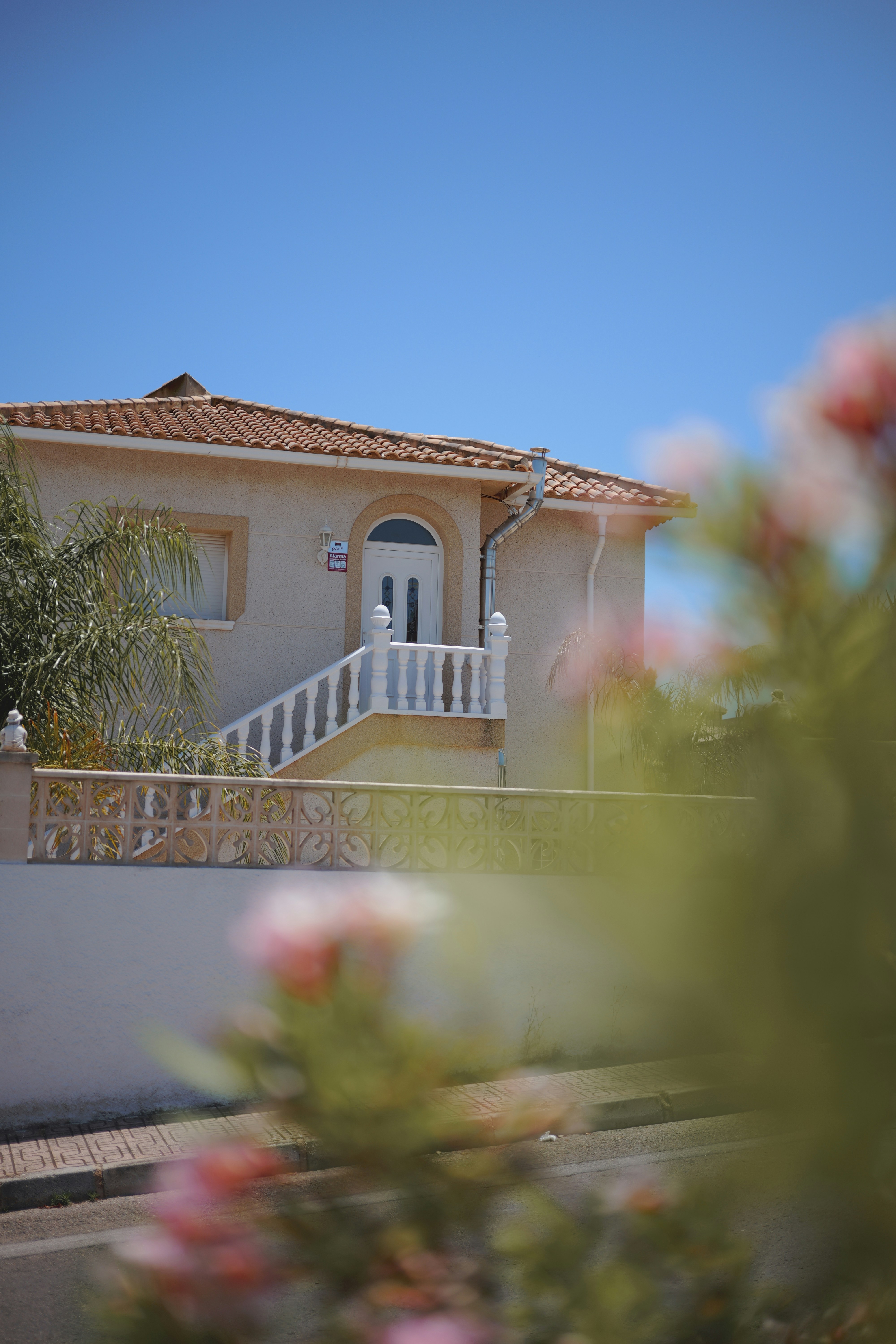 Sunlit Mediterranean-style house framed by a white staircase and a garden wall, with blurred foreground flowers creating depth.