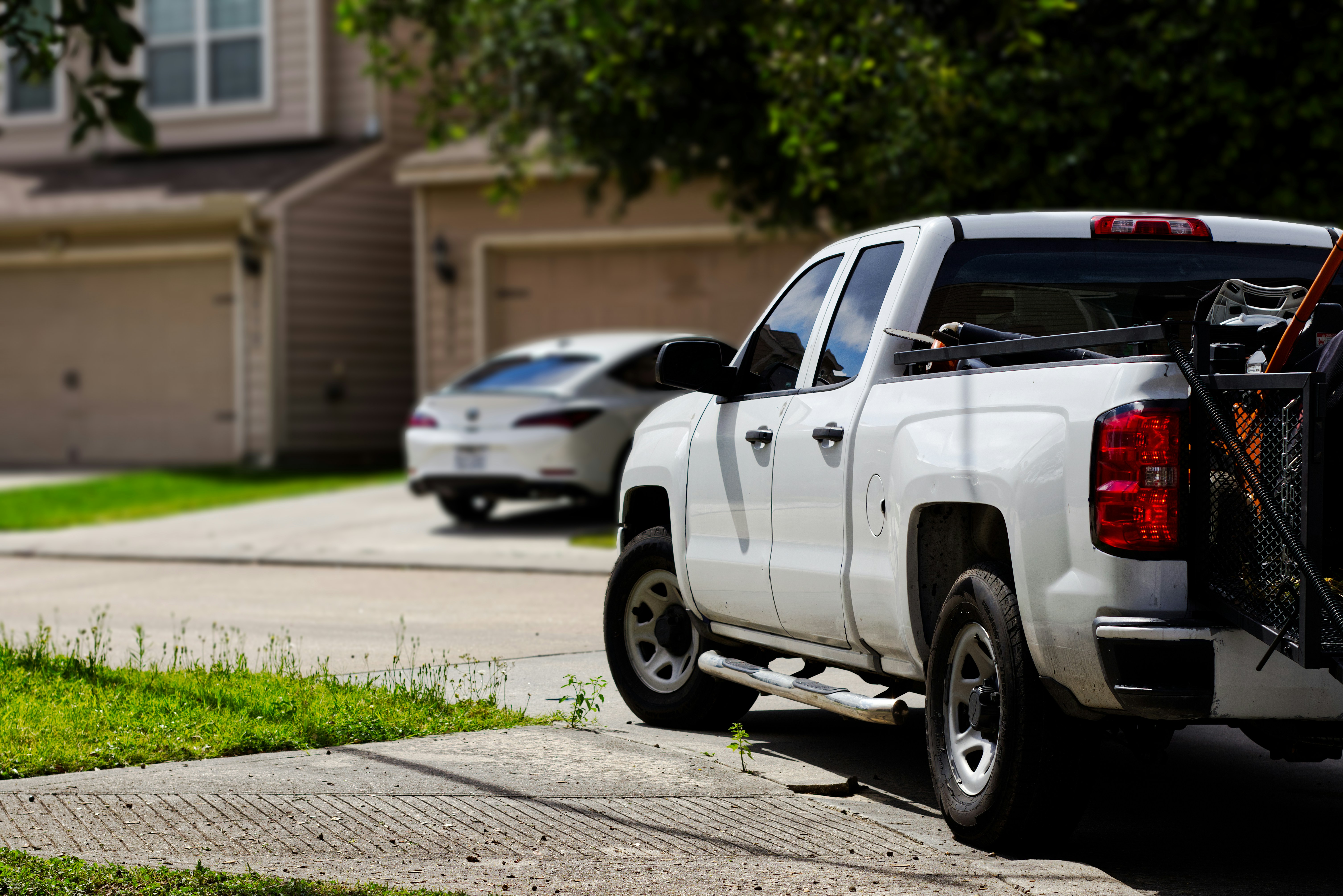 White pickup with a trailer bed gear parked in a suburban driveway; blurred background of a beige house and another car, with green lawn and sidewalk in foreground.