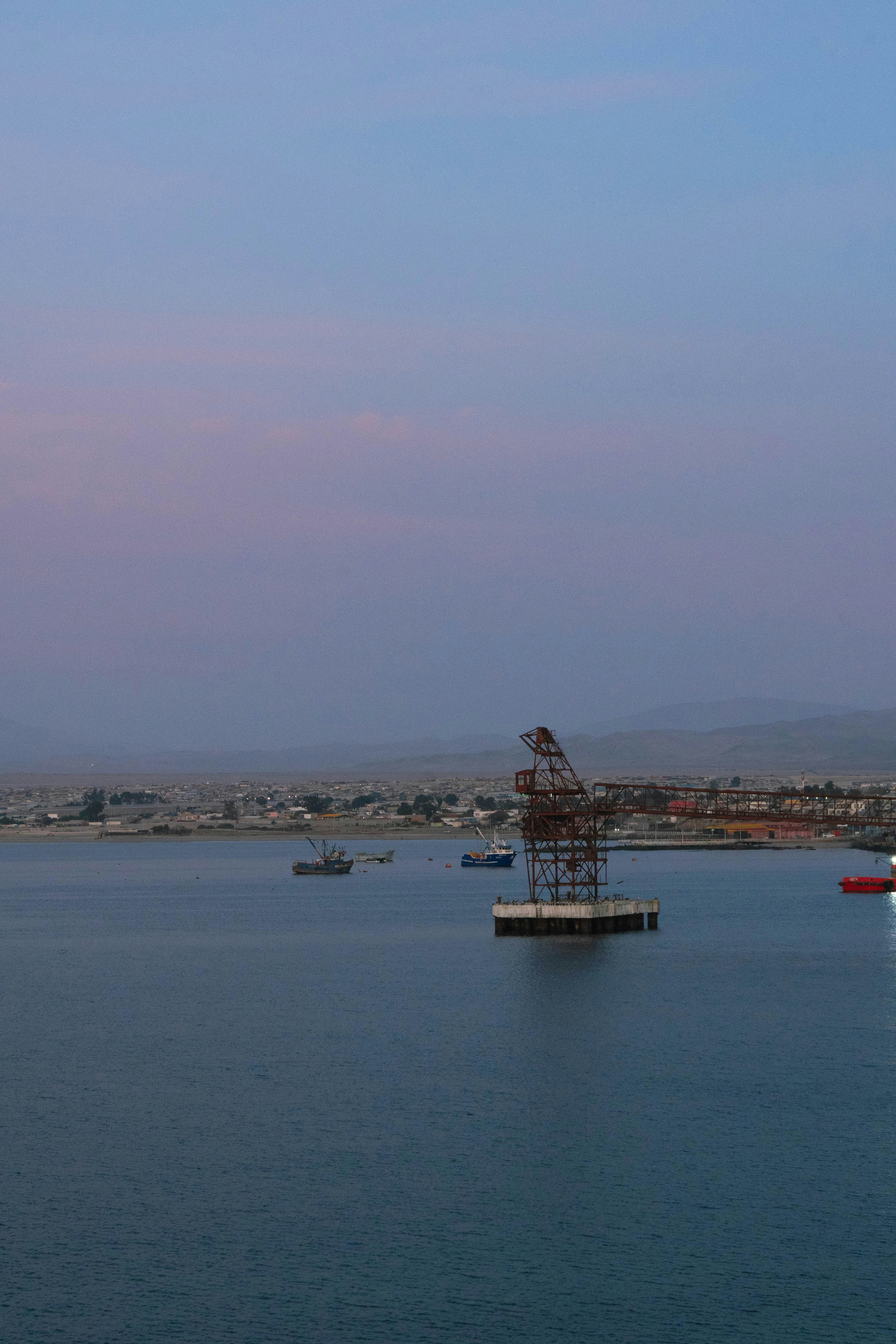 Harbor scene with calm water and distant land; a rusted crane-like structure rises from a small platform in the water, boats scattered along the horizon, pastel sky at dusk. This is a photographic capture, not a painting or illustration.