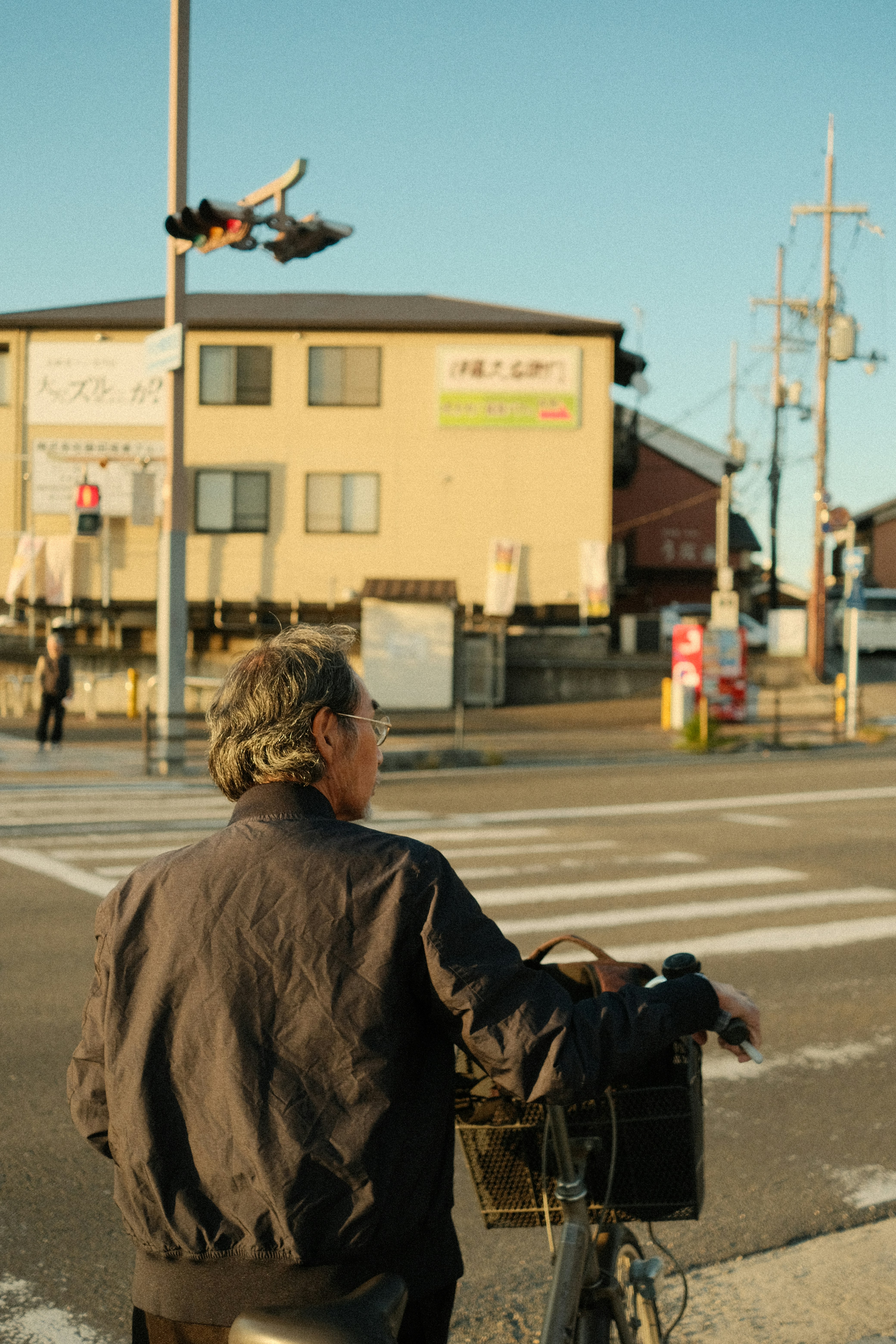 Elderly man with a bicycle stands at a crosswalk, back to camera, as a skateboarder soars above in the street under a clear morning sky.