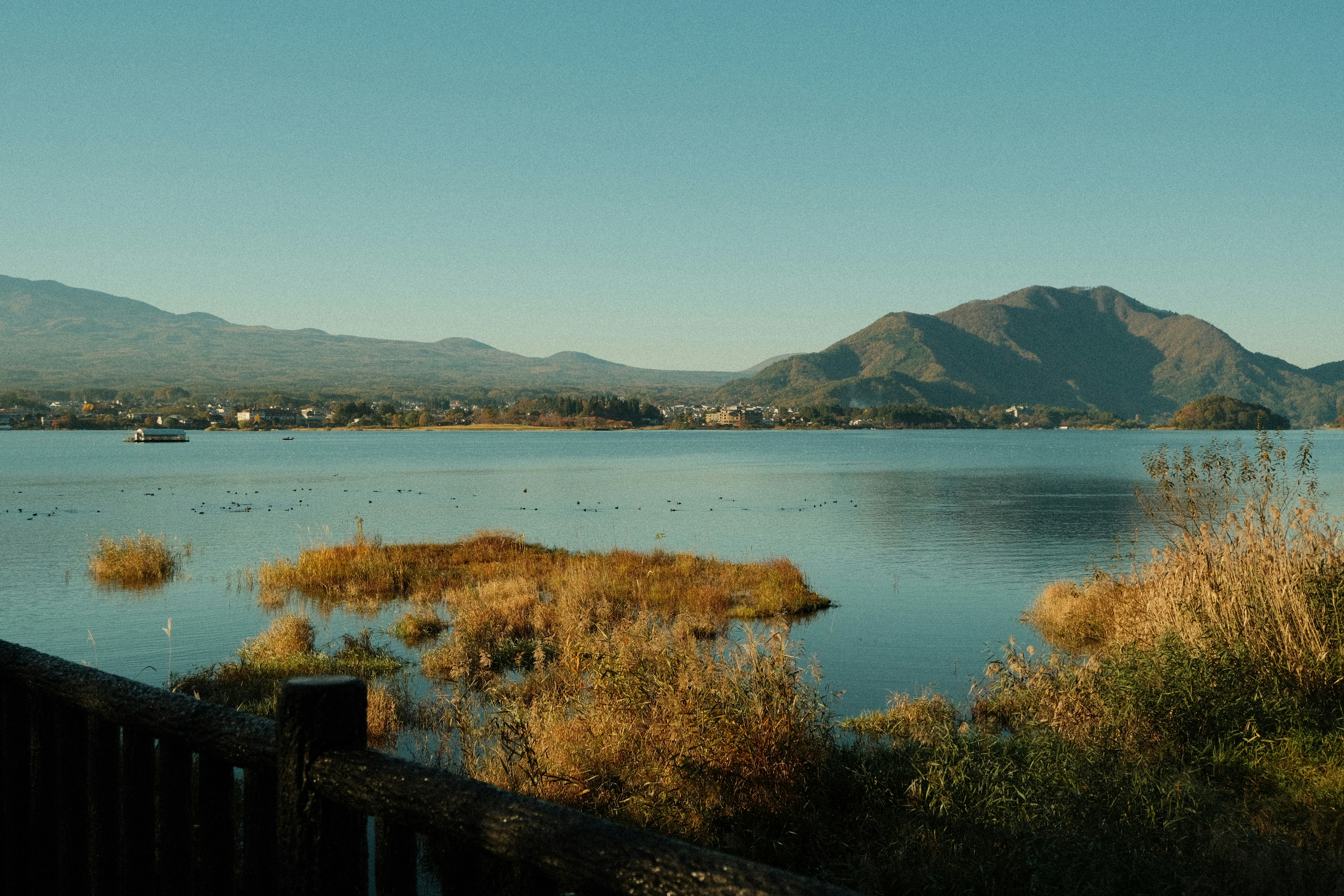Soft-hued lake with grassy islands and distant mountains under a clear sky, framed by a dark fence in the foreground. A tranquil landscape scene suitable for a travel or nature feature.